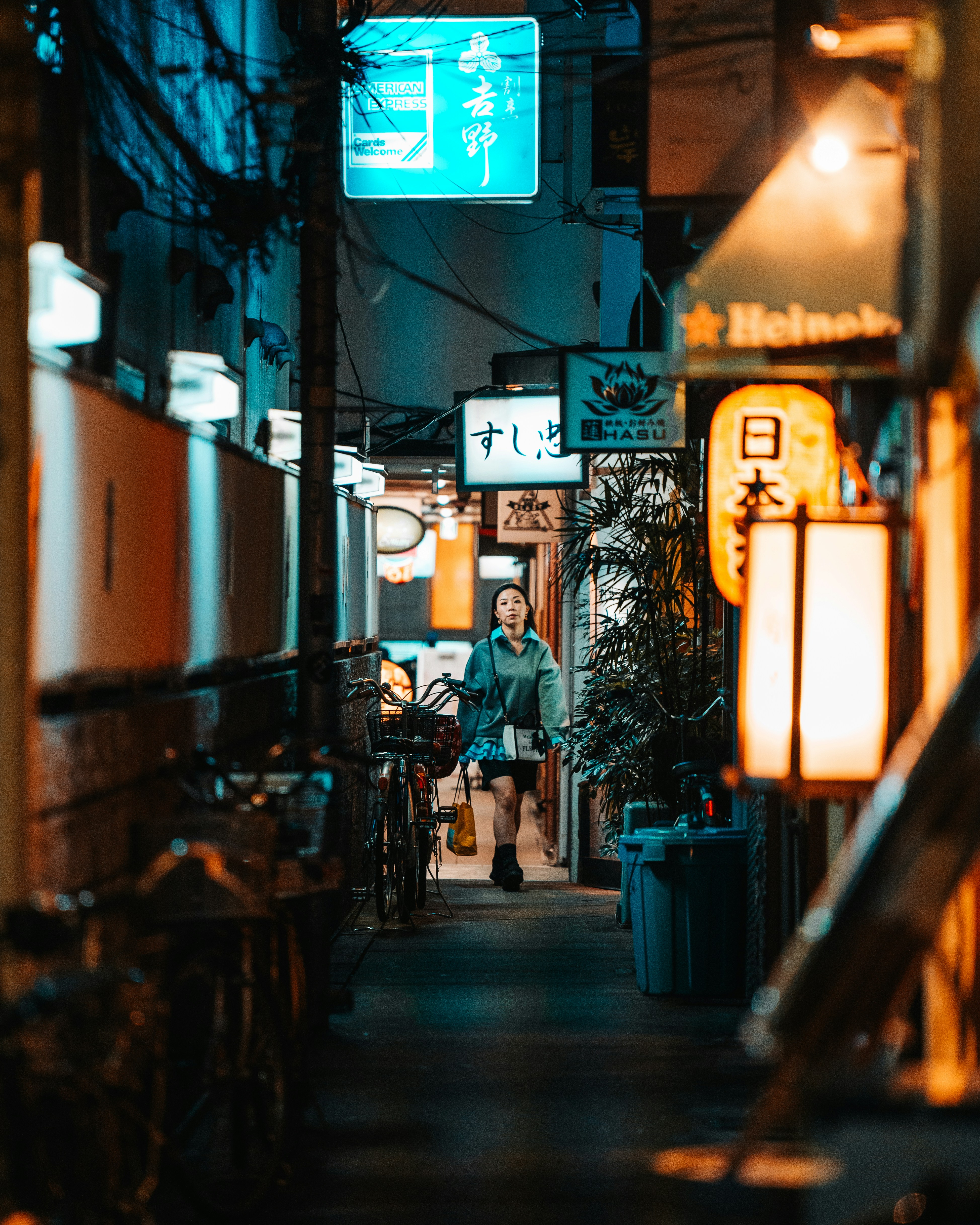 A young Japanese woman walks directly towards the camera, making eye contact on a narrow, traditional alley in Osaka at night. The scene is lit by soft lantern light and the glow of distant neon signs, captured with a telephoto lens to compress the urban atmosphere.