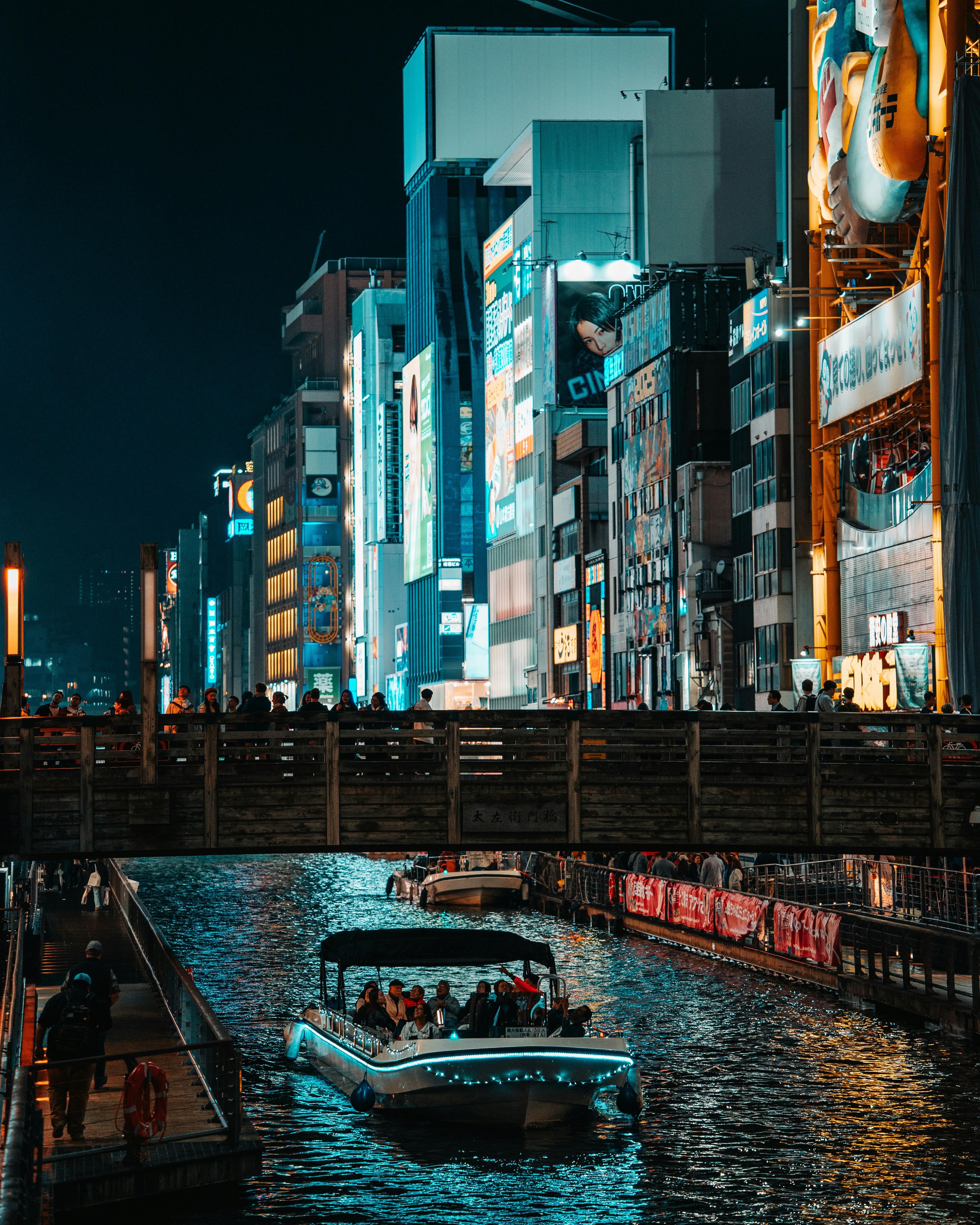 A dynamic night scene on the Dotonbori Canal in Osaka. A tourist boat with people glides under an illuminated bridge crowded with pedestrian silhouettes. Vibrant neon signs and city lights create picturesque reflections on the water, capturing the energy and atmosphere of the famous district.