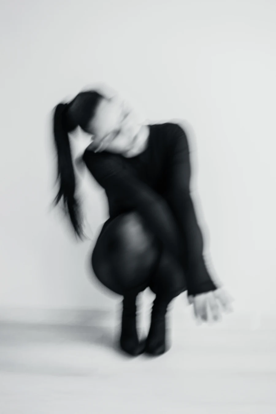 Monochrome long-exposure portrait of a crouched figure with a ponytail against a pale wall. Blur emphasizes pose and motion, with minimal background distraction.