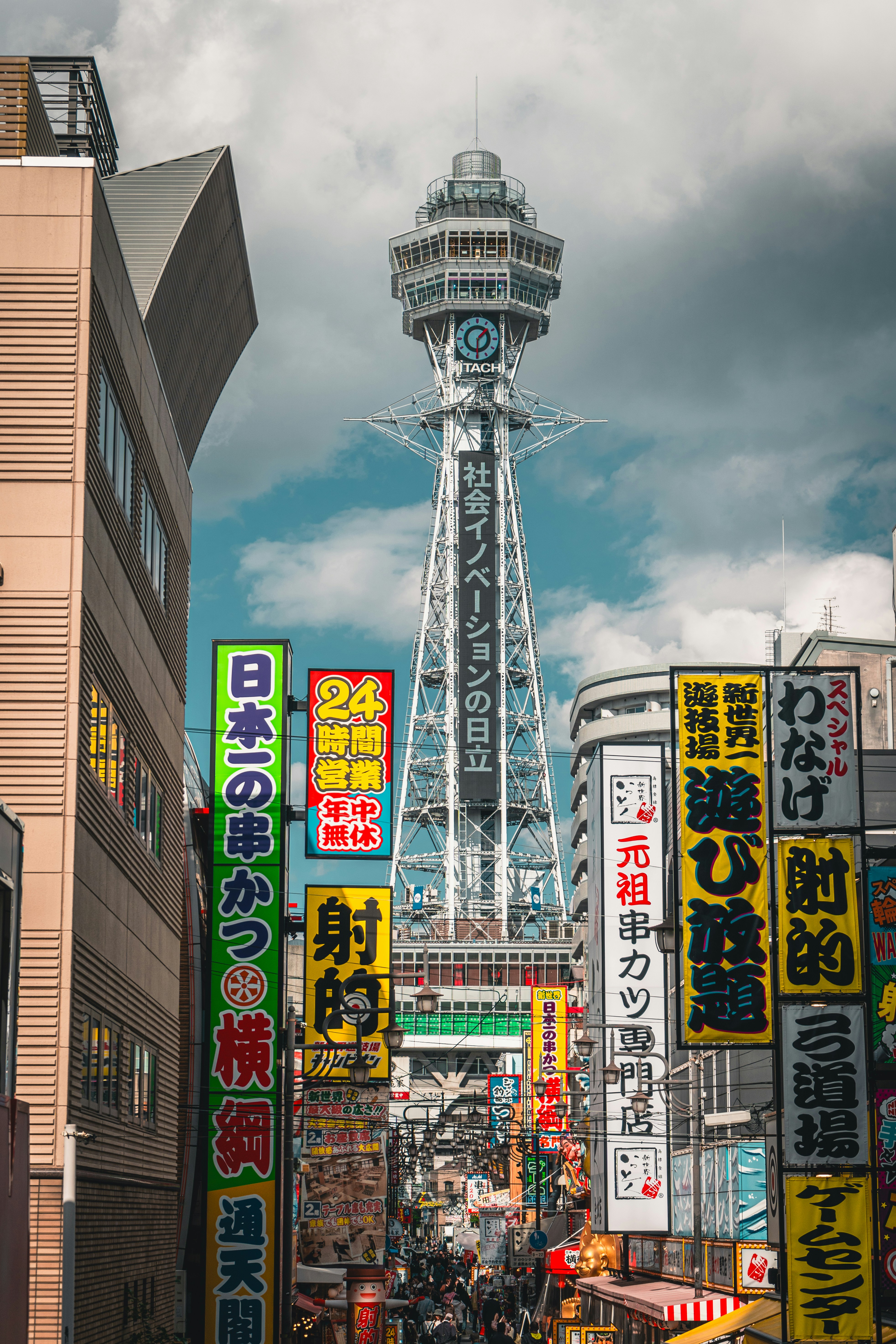 Tsutenkaku Tower captured from the bustling shopping street of Osaka's Shinsekai district. The frame captures the perspective of the vibrant street with colorful signs, leading the eye directly to the famous tower - symbol of this colorful historic district.