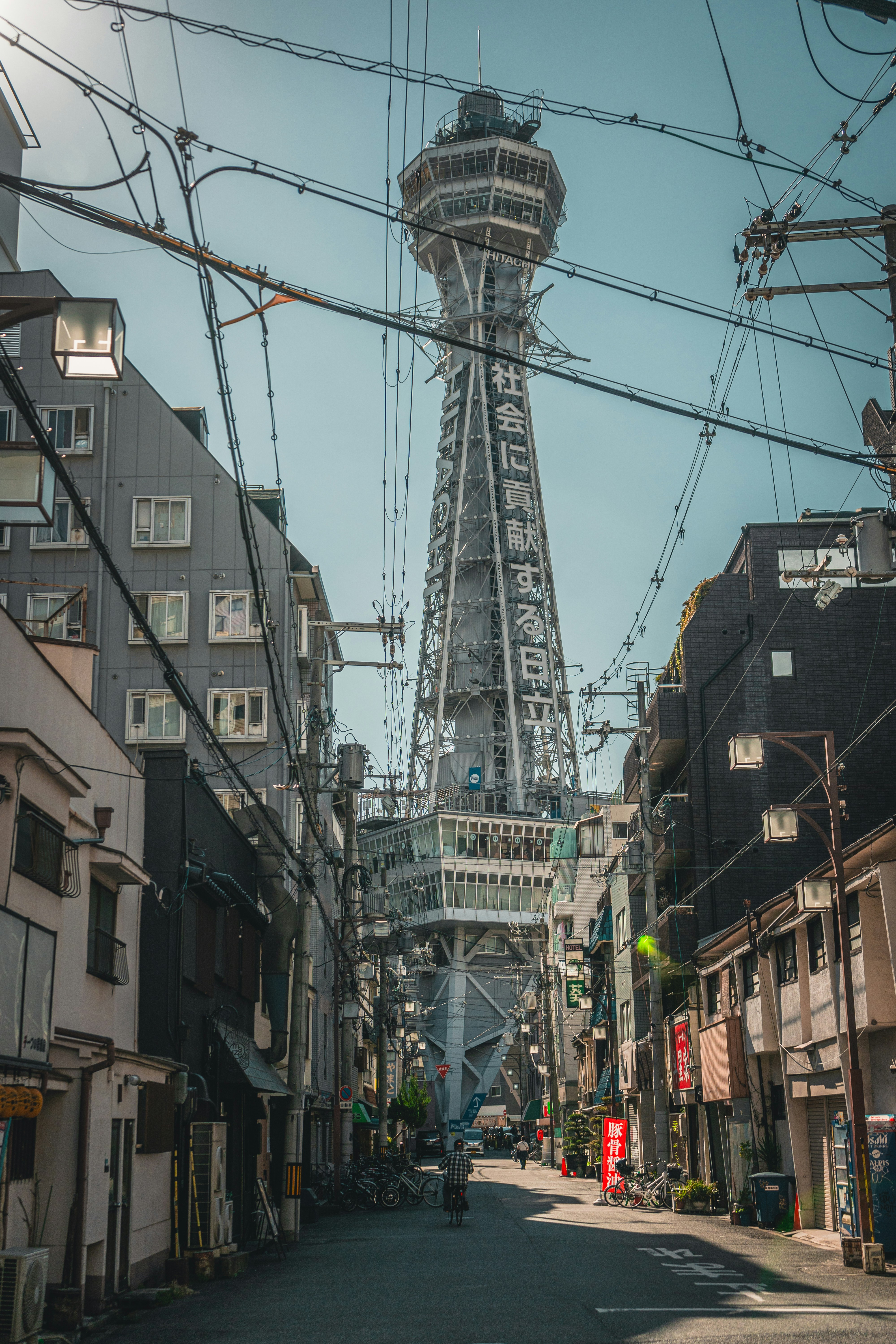 Tsutenkaku Tower captured from the bustling shopping street of Osaka's Shinsekai district. The frame captures the perspective of the vibrant street with colorful signs, leading the eye directly to the famous tower - symbol of this colorful historic district.