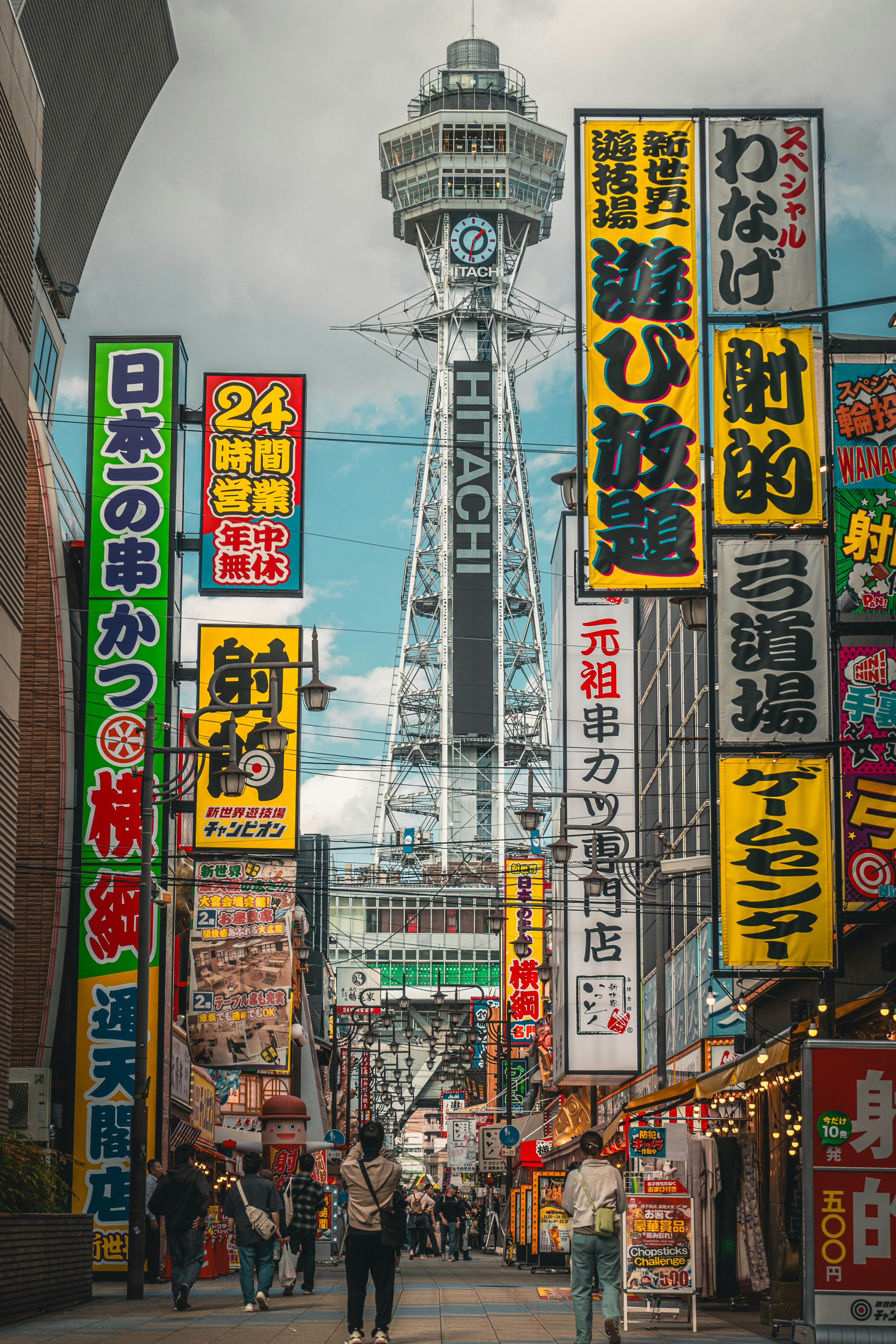Tsutenkaku Tower captured from the bustling shopping street of Osaka's Shinsekai district. The frame captures the perspective of the vibrant street with colorful signs, leading the eye directly to the famous tower - symbol of this colorful historic district.