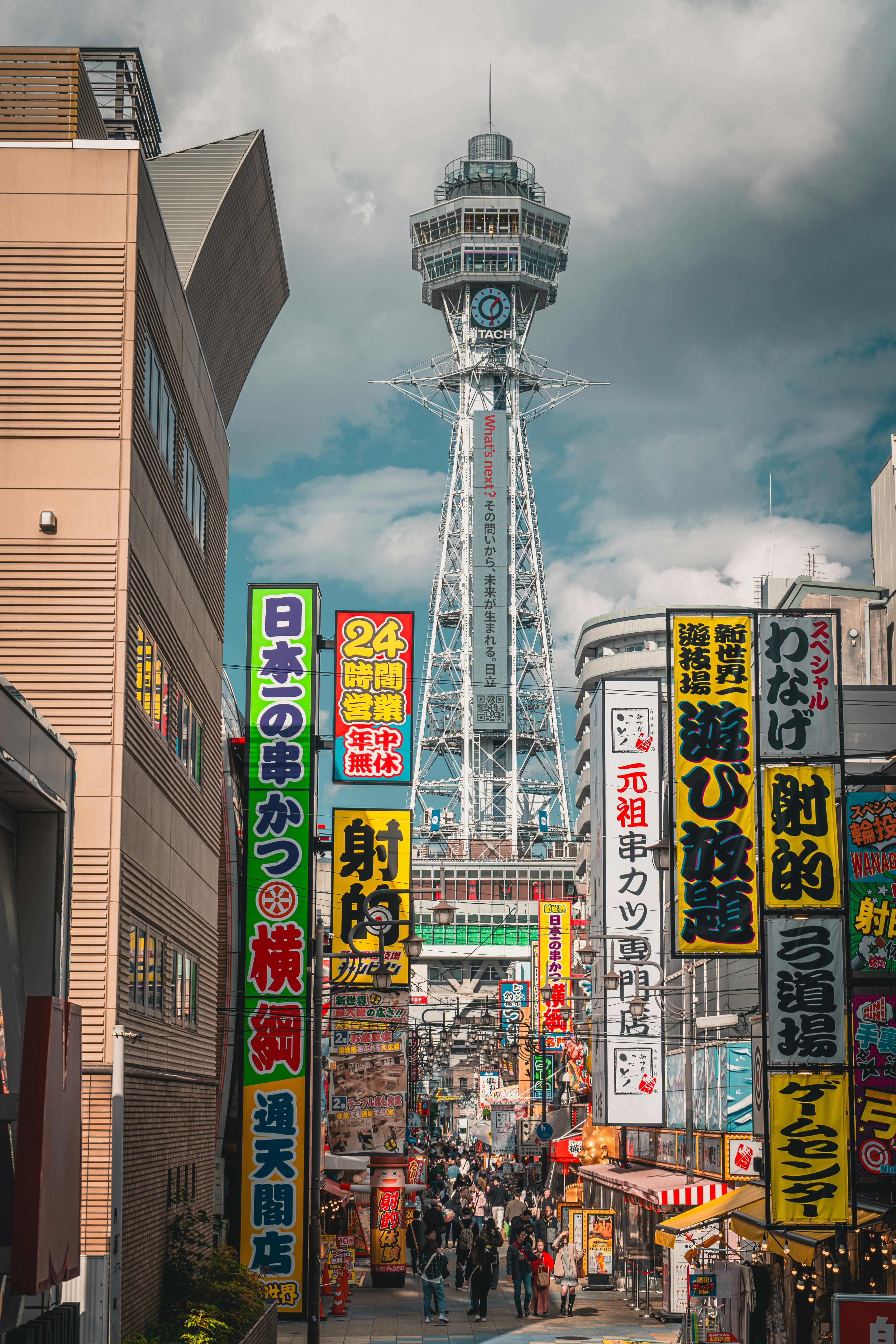 Tsutenkaku Tower captured from the bustling shopping street of Osaka's Shinsekai district. The frame captures the perspective of the vibrant street with colorful signs, leading the eye directly to the famous tower - symbol of this colorful historic district.