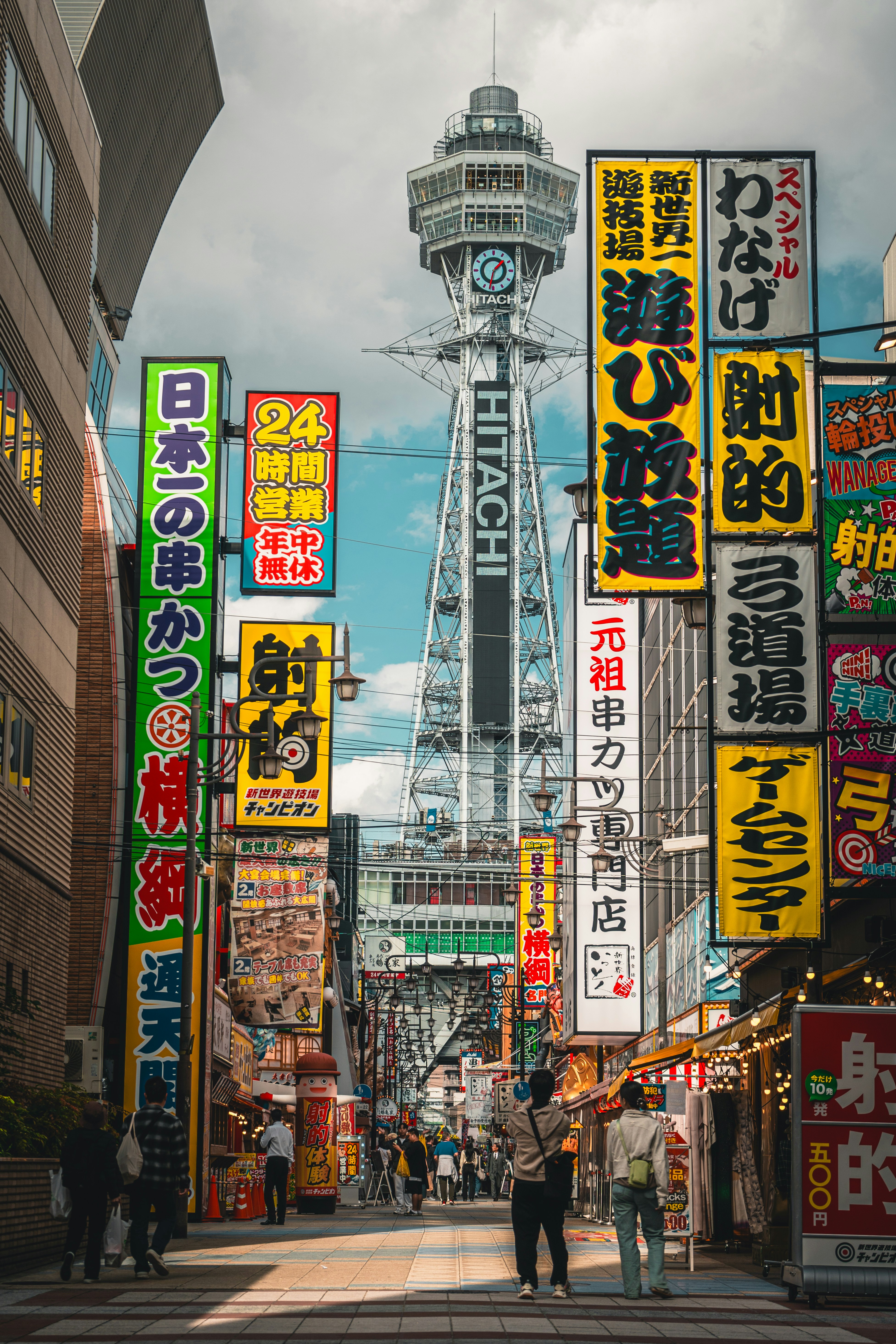 Tsutenkaku Tower captured from the bustling shopping street of Osaka's Shinsekai district. The frame captures the perspective of the vibrant street with colorful signs, leading the eye directly to the famous tower - symbol of this colorful historic district.