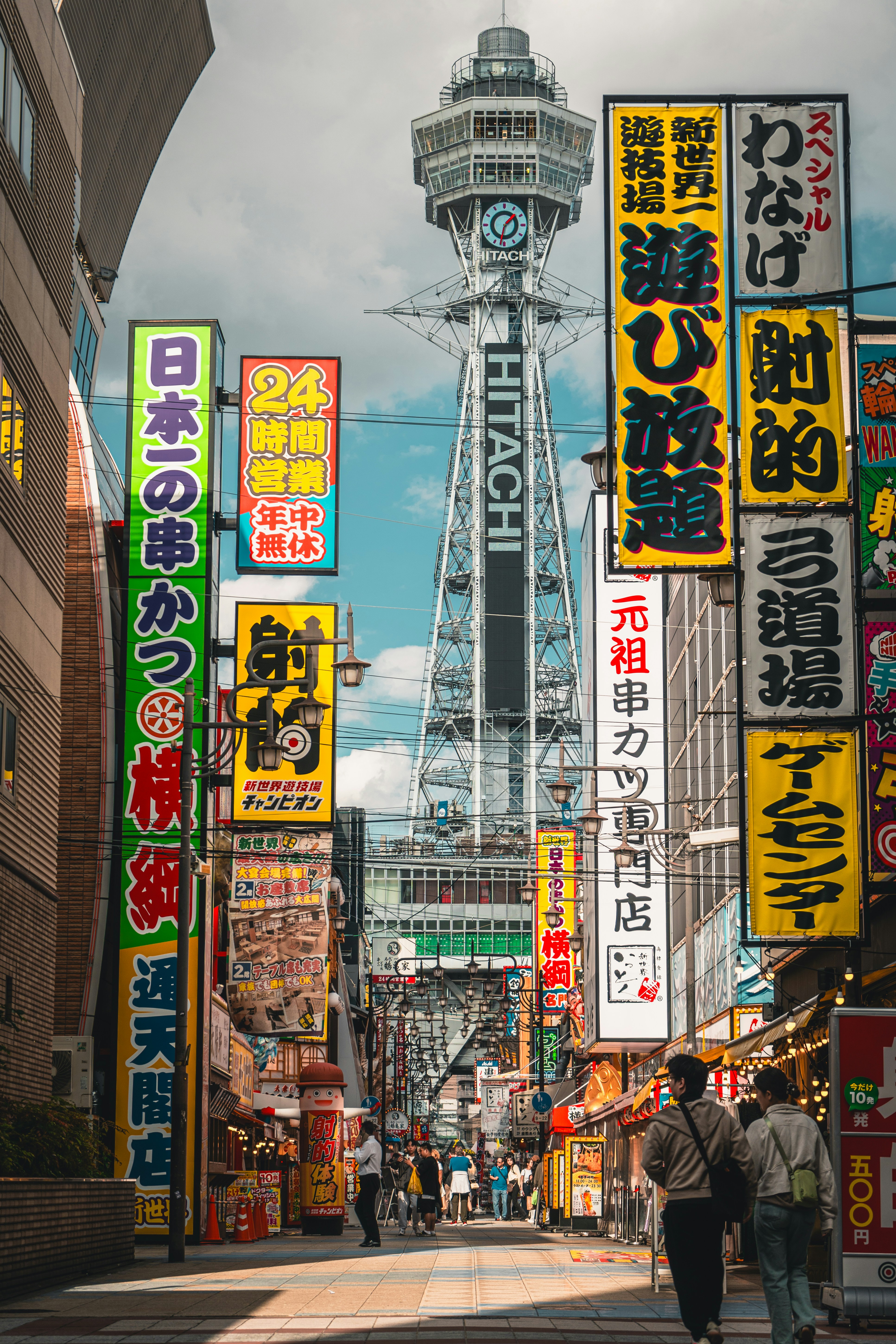 Tsutenkaku Tower captured from the bustling shopping street of Osaka's Shinsekai district. The frame captures the perspective of the vibrant street with colorful signs, leading the eye directly to the famous tower - symbol of this colorful historic district.