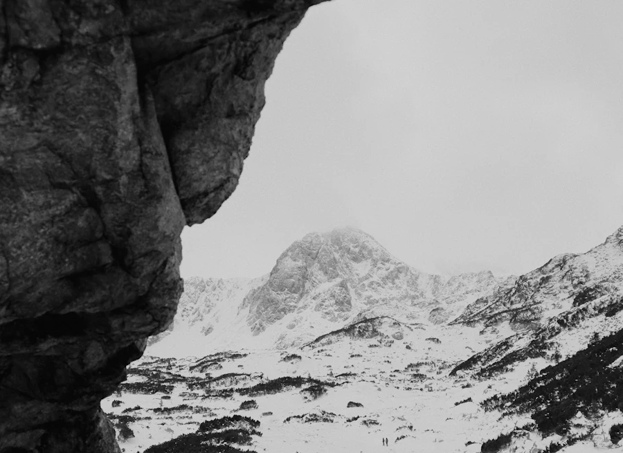 Monochrome alpine landscape framed by a dark rock on the left, with a snow-covered ridge and distant peak. The composition emphasizes texture and contrast against the rugged foreground.