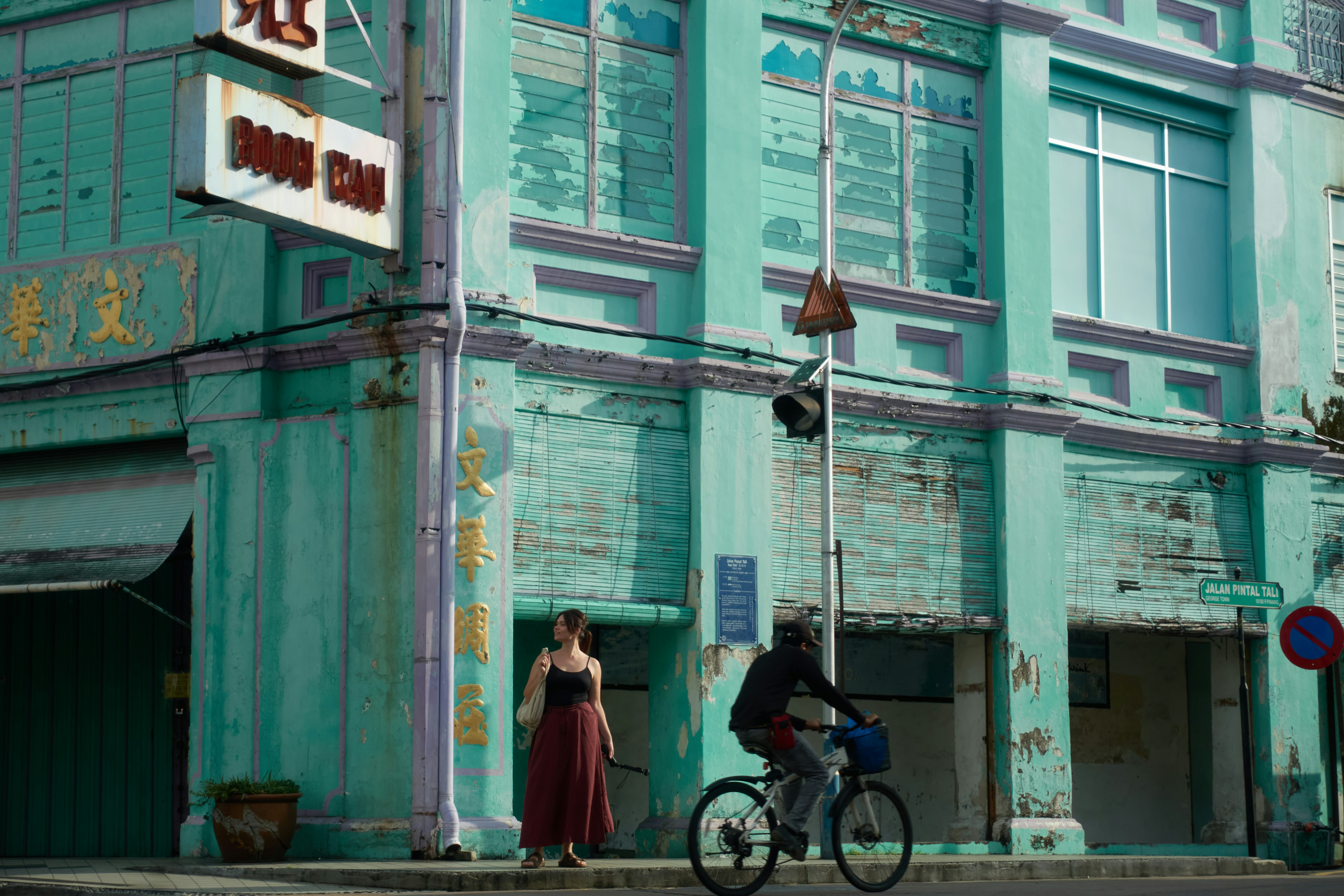 Teal Facade, Quiet Moment: A Woman and Cyclist in a Weathered Street
