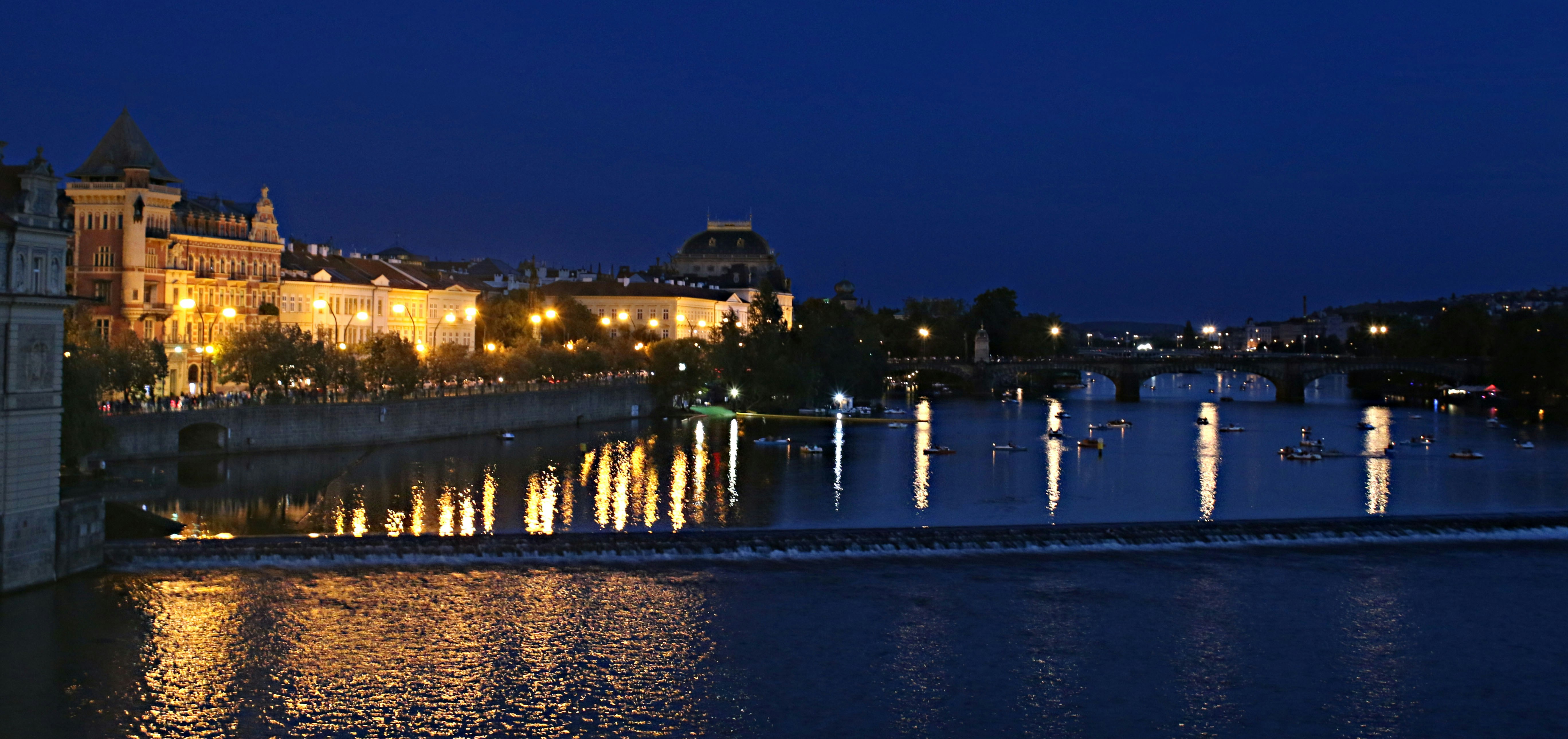 The nighttime magic of Prague is mirrored on the calm waters of the Vltava River. The lights of historic buildings brighten the riverside, while small boats glide under the deep blue sky, creating a romantic and vibrant atmosphere in the heart of the city.
