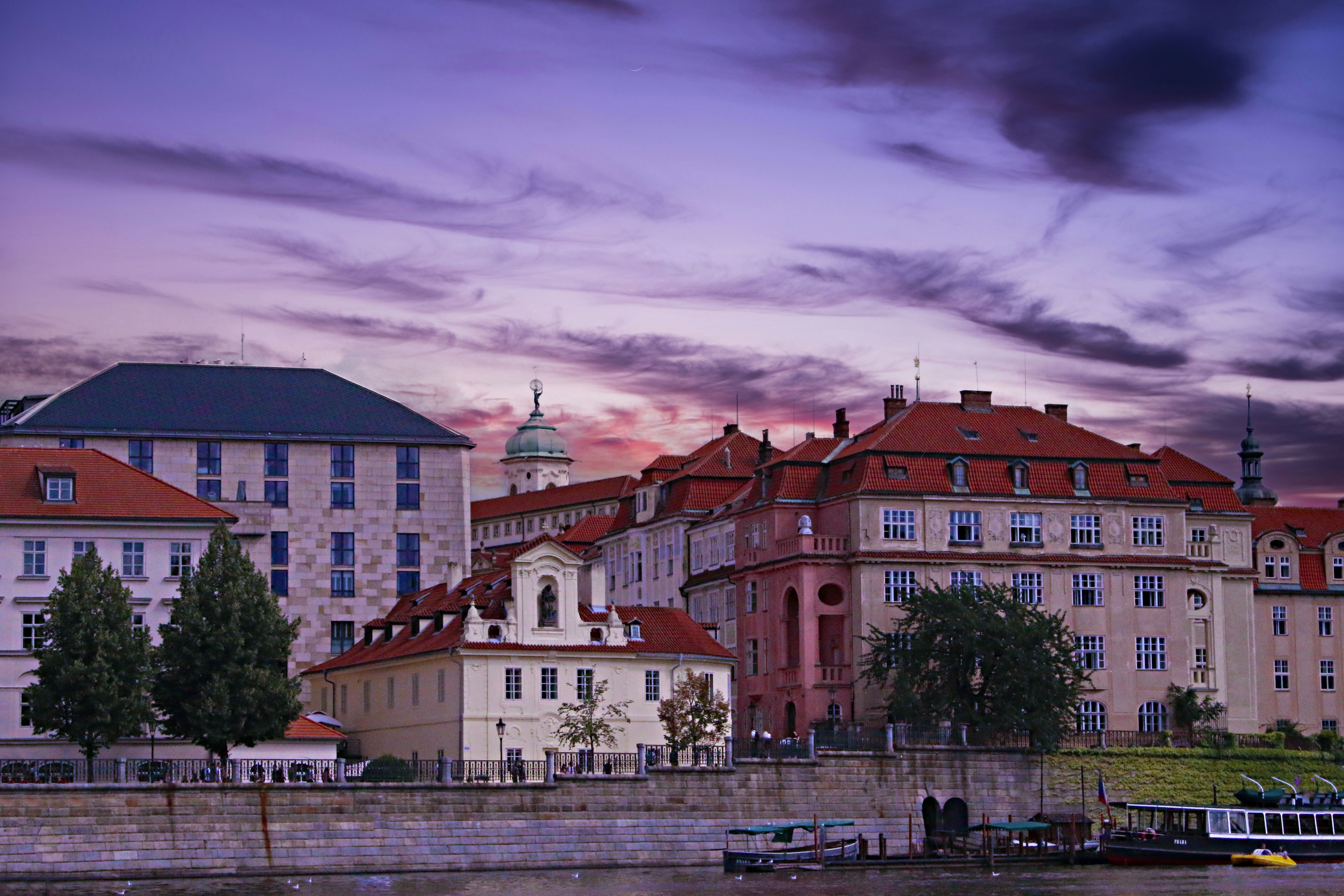 The purple and pink sunset sky blends with Prague’s architecture, where the red-roofed buildings stand gracefully along the serene Vltava River. A scene that unites the natural beauty of twilight with the historic charm of the city.