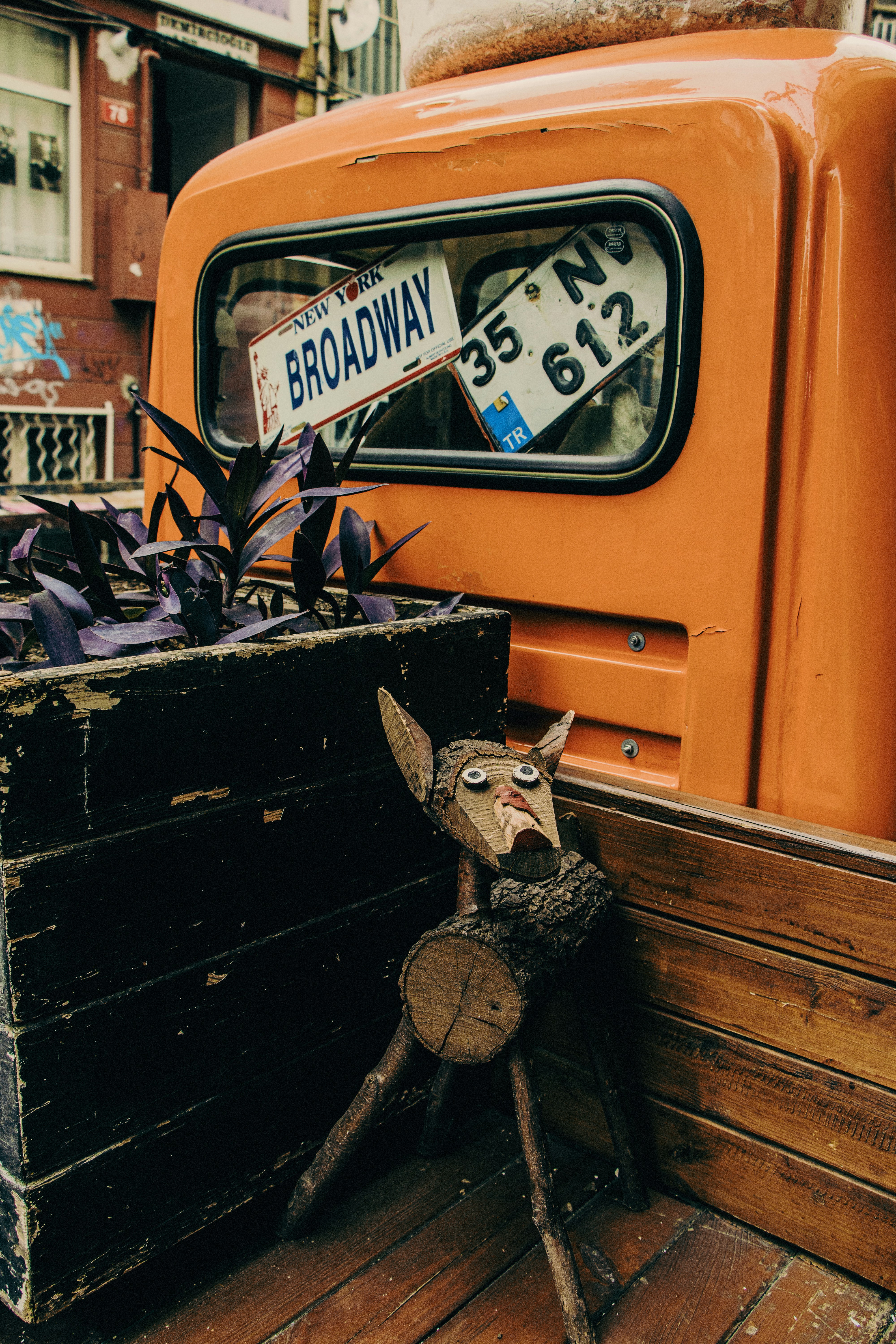 Rust and Route: A Wooden Dog Guards a Planter by an Orange Vintage Truck