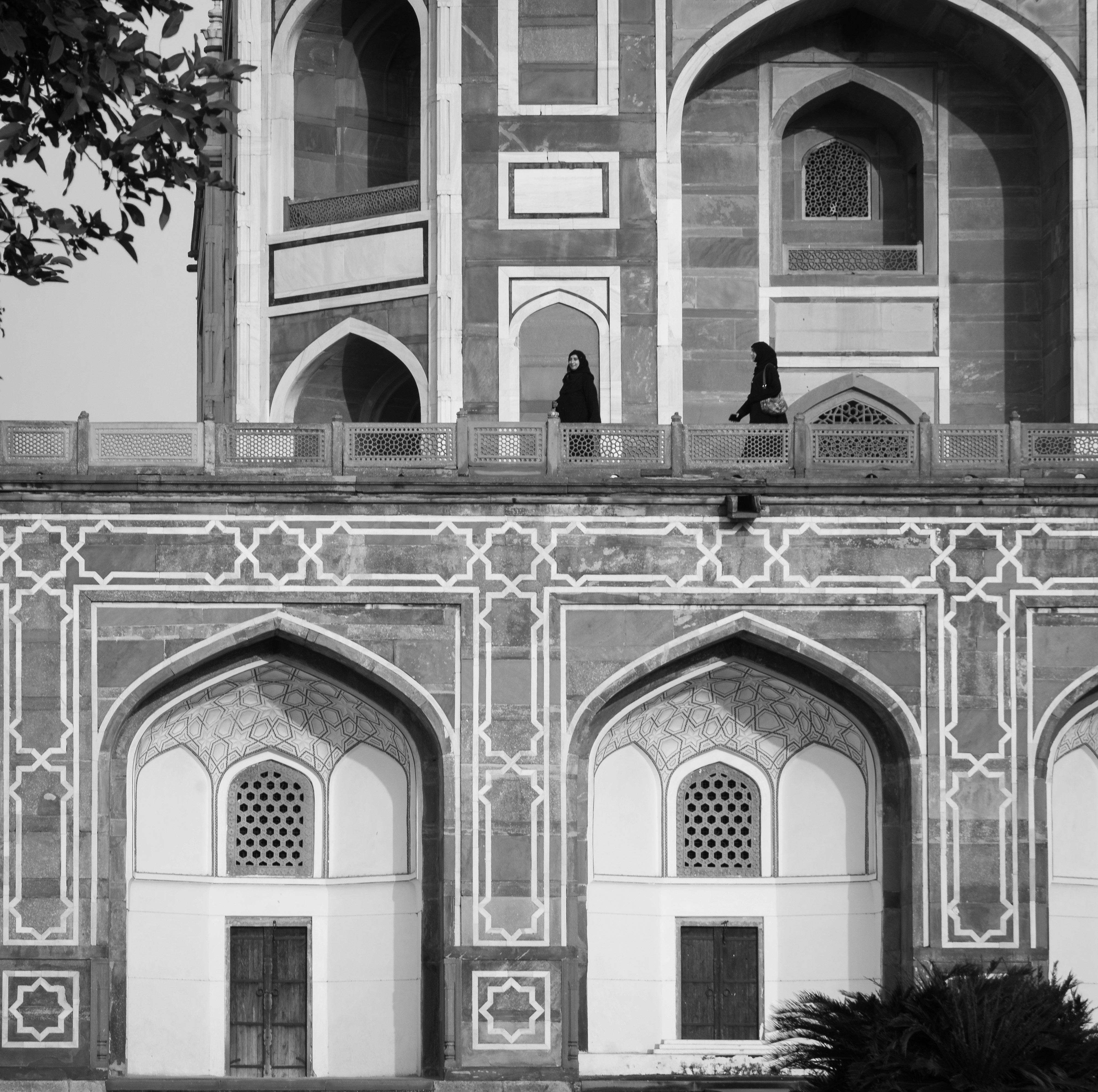 This photograph captures the multi-tiered facade of Humayun's Tomb in Delhi, emphasizing its Mughal architectural symmetry and use of red sandstone with white marble inlay. Two women dressed in black abayas are visible walking along this upper balcony, providing a sense of scale and human activity against the backdrop of the large arched niches and windows. This image is excellent for content related to Islamic architecture, Indian heritage, cultural dress, and the geometry and scale of historic monuments.