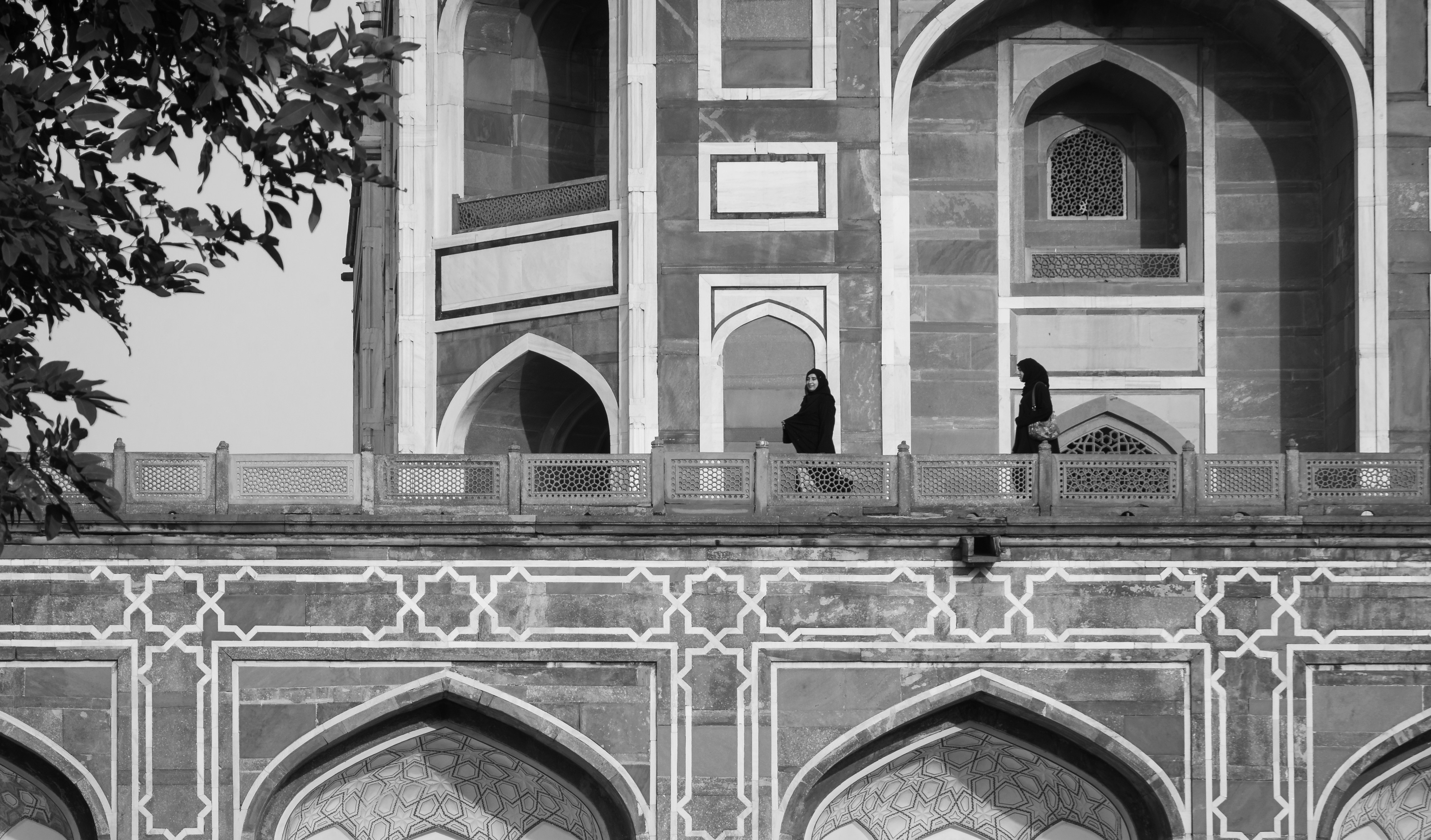 This photograph captures the multi-tiered facade of Humayun's Tomb in Delhi, emphasizing its Mughal architectural symmetry and use of red sandstone with white marble inlay. Two women dressed in black abayas are visible walking along this upper balcony, providing a sense of scale and human activity against the backdrop of the large arched niches and windows. This image is excellent for content related to Islamic architecture, Indian heritage, cultural dress, and the geometry and scale of historic monuments.