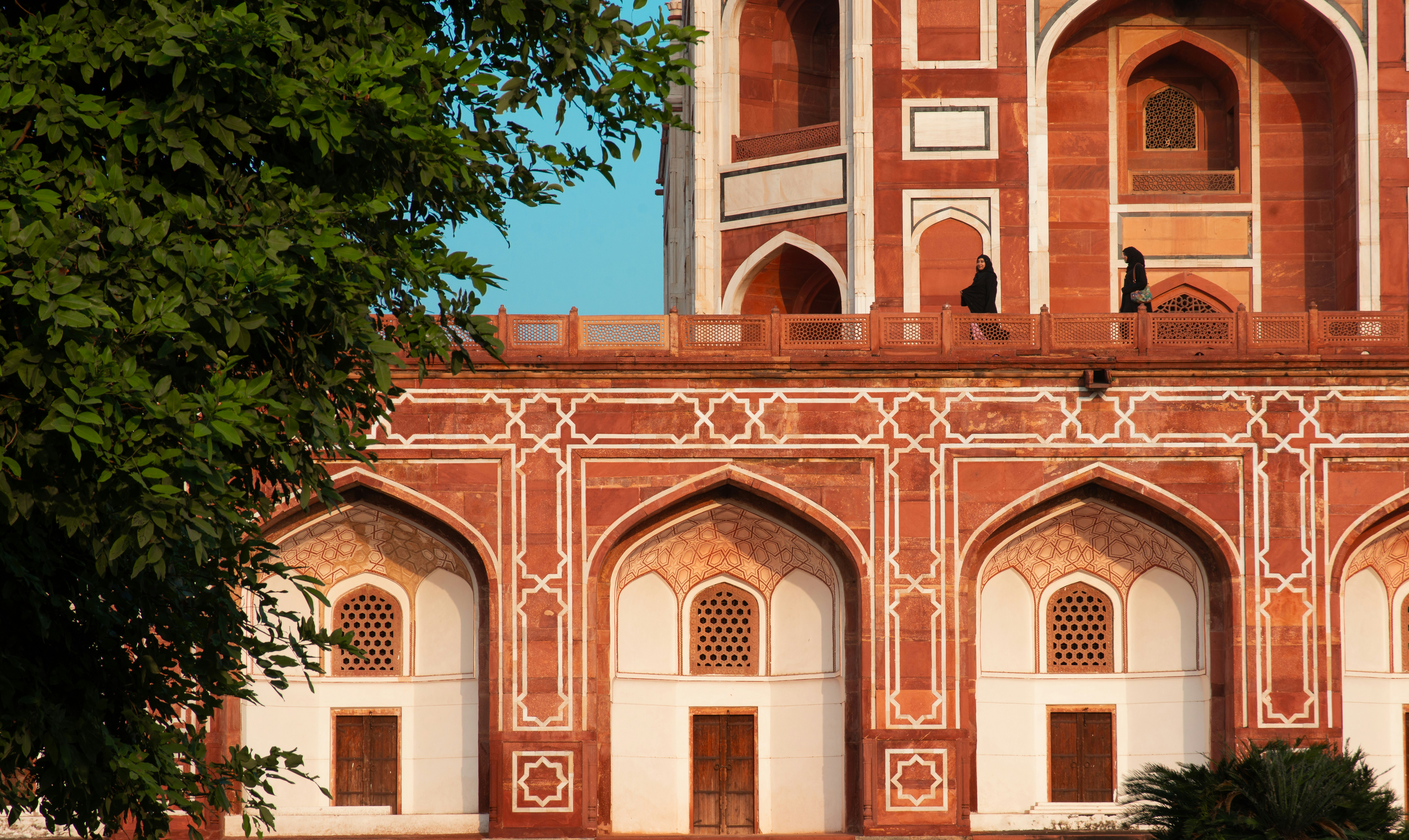 This photograph captures the multi-tiered facade of Humayun's Tomb in Delhi, emphasizing its Mughal architectural symmetry and use of red sandstone with white marble inlay. Two women dressed in black abayas are visible walking along this upper balcony, providing a sense of scale and human activity against the backdrop of the large arched niches and windows. This image is excellent for content related to Islamic architecture, Indian heritage, cultural dress, and the geometry and scale of historic monuments.