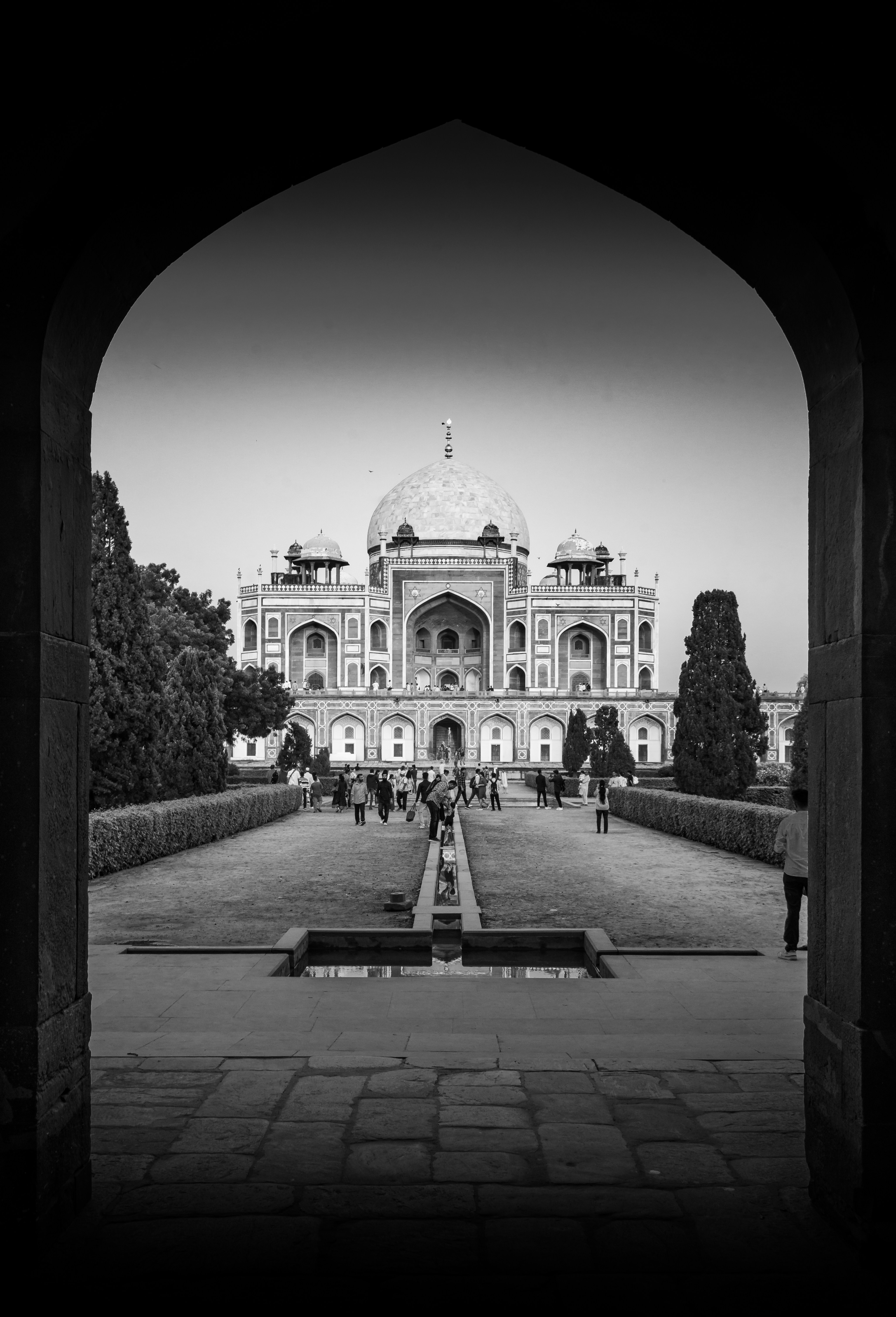 The symmetrical, monumental Humayun's Tomb in Delhi framed by a dark foreground archway. The structure's iconic Mughal architecture is highlighted by the high contrast, emphasizing the white marble dome and inlays against the red sandstone facade. The Charbagh garden layout is visible in the foreground, with a central water channel and manicured hedges leading the eye toward the tomb. Several visitors are walking on the paths, providing a sense of scale. This image is suitable for content related to historic Indian architecture, Mughal garden design, fine art photography, and monumental scale.