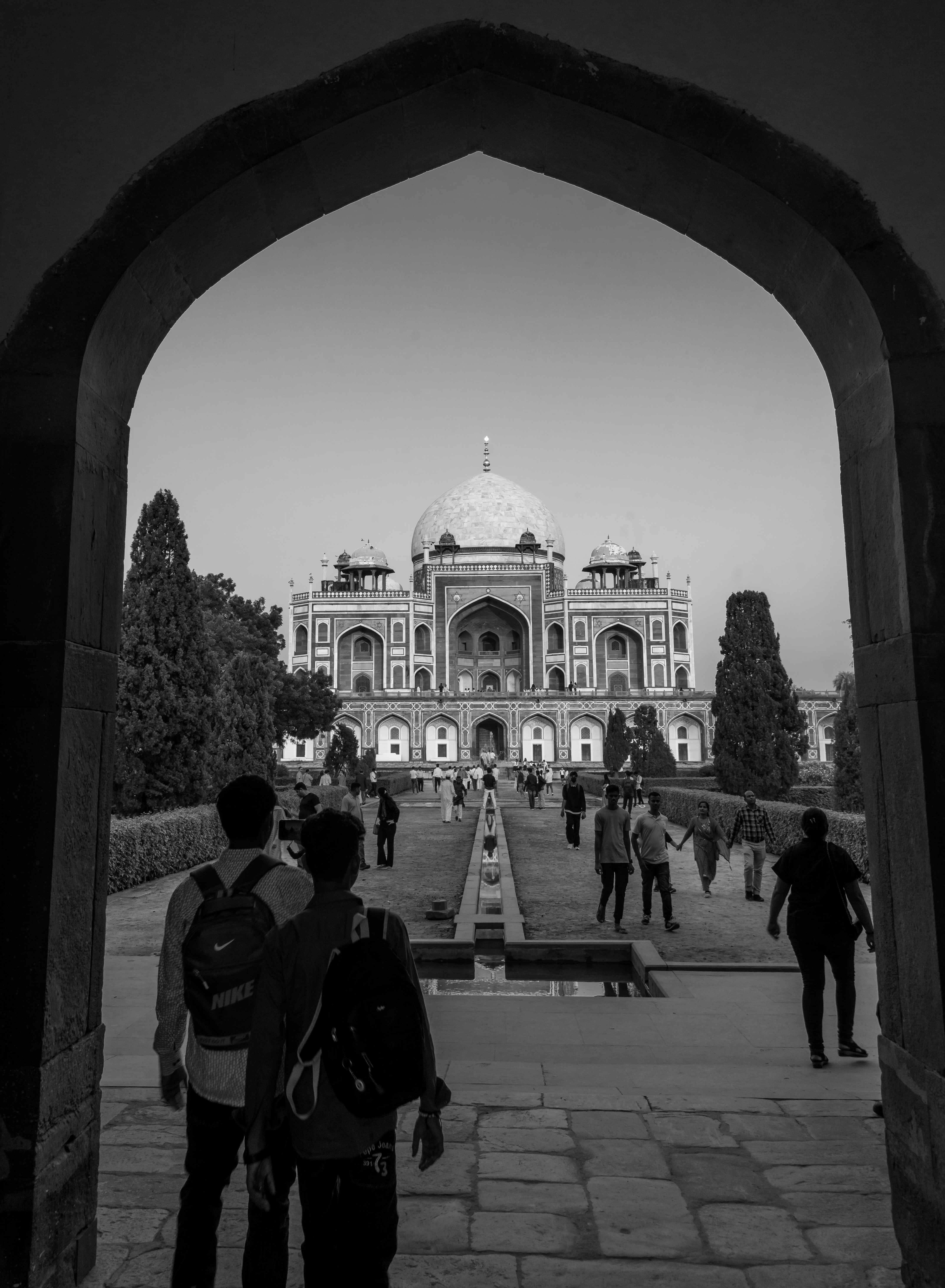 The symmetrical, monumental Humayun's Tomb in Delhi framed by a dark foreground archway. The structure's iconic Mughal architecture is highlighted by the high contrast, emphasizing the white marble dome and inlays against the red sandstone facade. The Charbagh garden layout is visible in the foreground, with a central water channel and manicured hedges leading the eye toward the tomb. Several visitors are walking on the paths, providing a sense of scale. This image is suitable for content related to historic Indian architecture, Mughal garden design, fine art photography, and monumental scale.