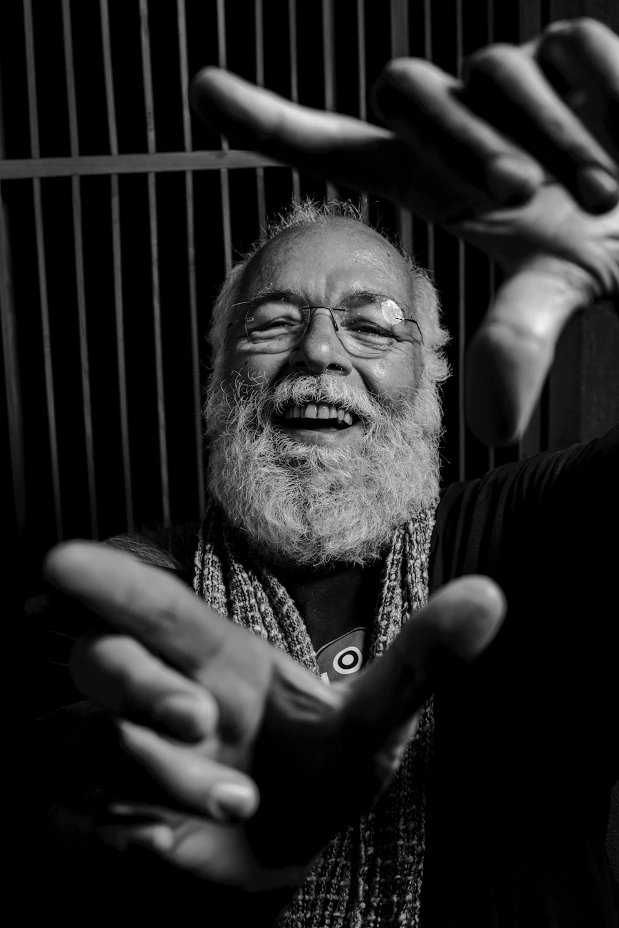Monochrome candid portrait of an elderly man with glasses and a white beard, smiling as hands reach toward the camera.
