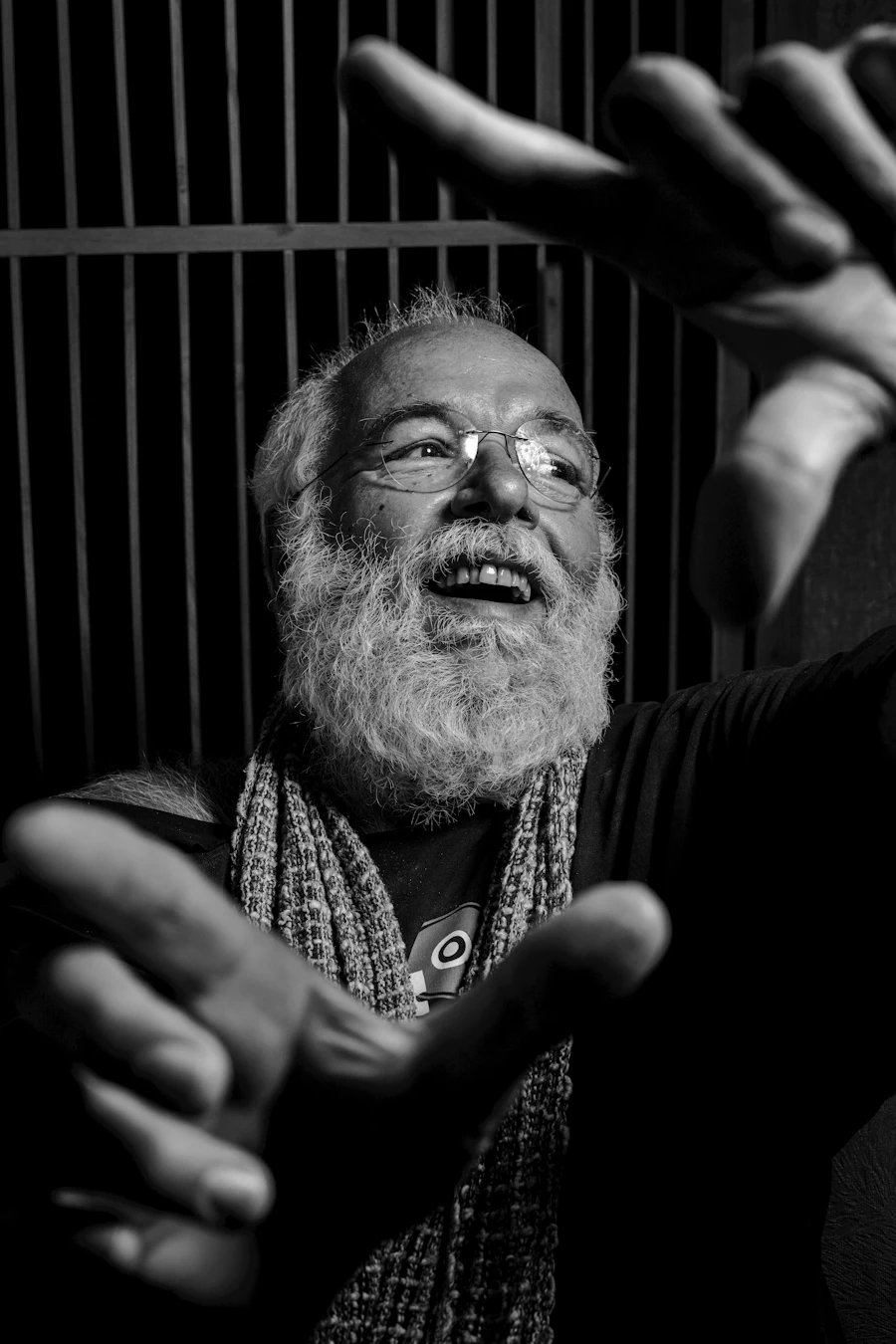 Black-and-white portrait of a bearded man with glasses reaching toward the camera, smiling broadly. Foreground hands blur while his face remains sharp.