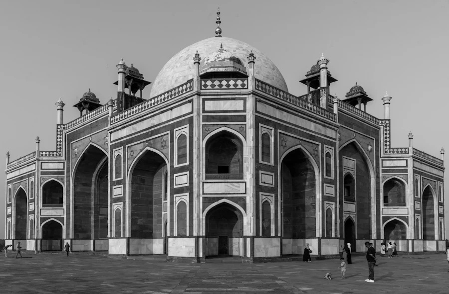 Black-and-white photograph of a Mughal tomb with a central dome and arched facades, set in a wide plaza. A few people provide scale against the monumental stonework.