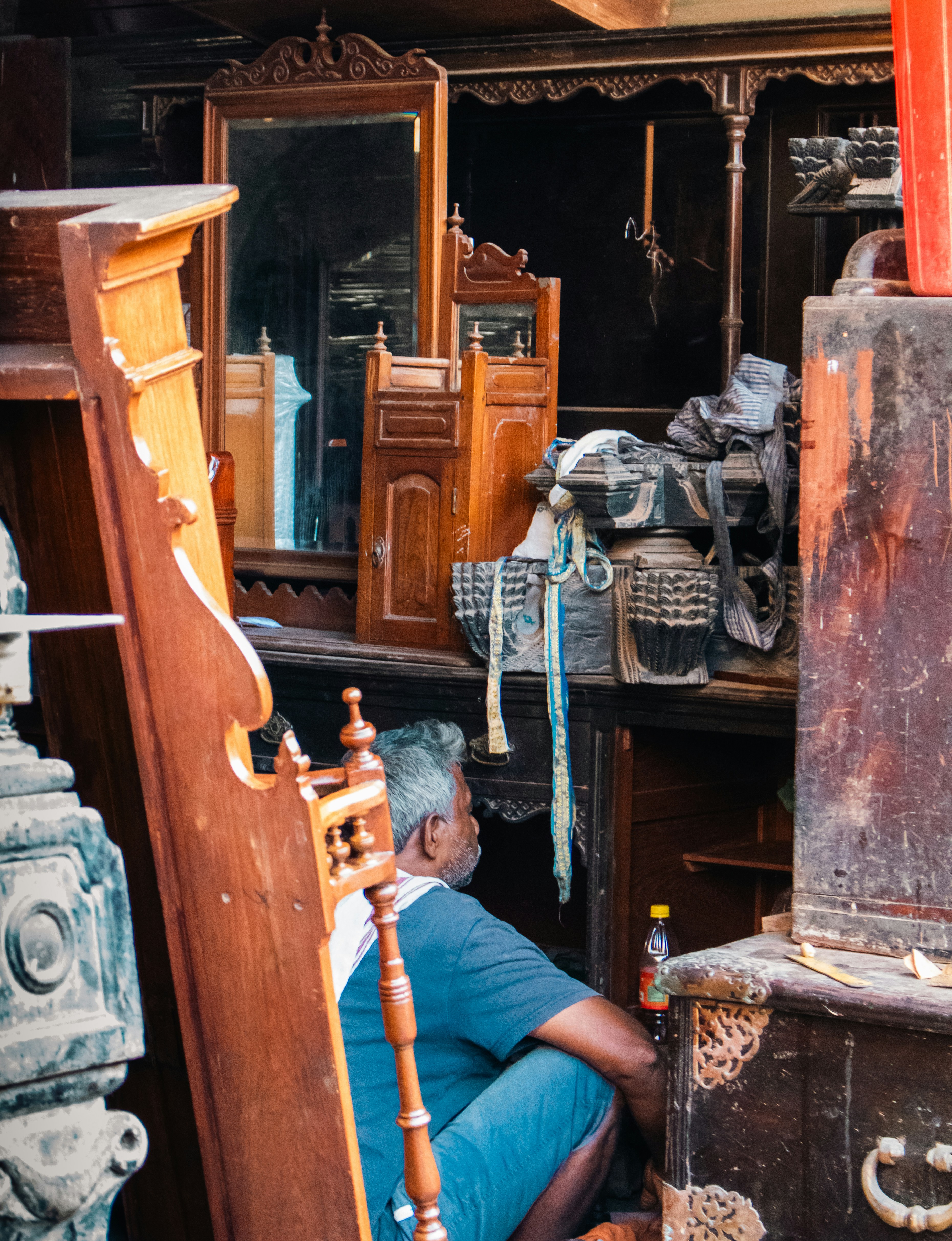 The cluttered interior of a secondhand furniture shop in Mumbai. A middle-aged man with grey hair, wearing a dark blue shirt and shorts, is sitting on the floor, partially obscuring the foreground. He is surrounded by an accumulation of dark, carved wooden furniture, including antique dressers, mirrors, and decorative pieces. The setting emphasizes the dense, layered collection of goods and the intricacy of Indian wooden craftsmanship. This image is suitable for content on vintage furniture, secondhand markets, wood carving, local commerce, or street trade in India.