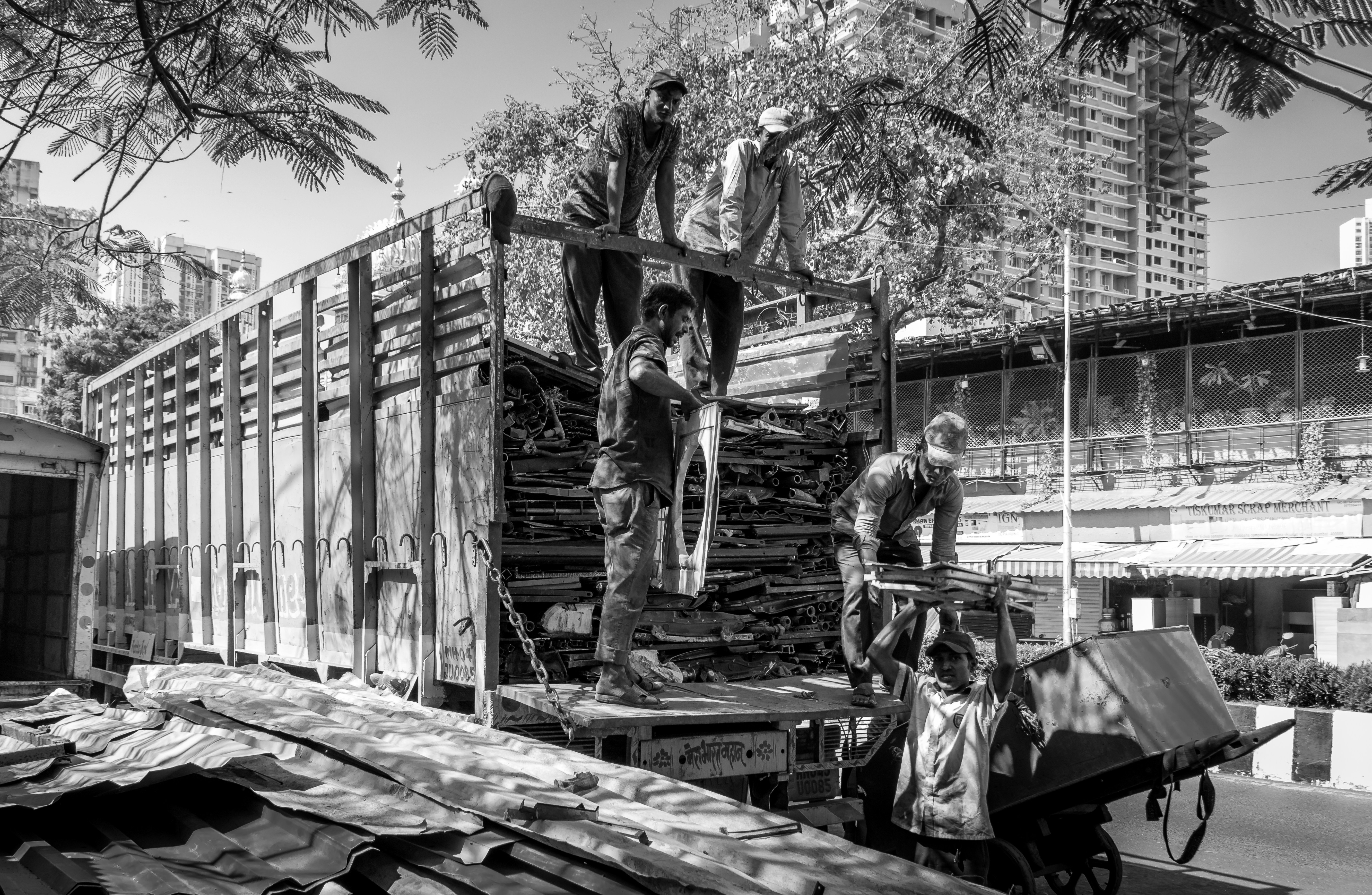 Four male laborers are engaged in the process of unloading flat sheets from the bed of a truck. The scene is shot from a low angle, emphasizing the physical labor and the height of the vehicle, characteristic of daily commerce and goods transport in a busy urban area like Mumbai. This image is suitable for content on manual labor and construction trade.