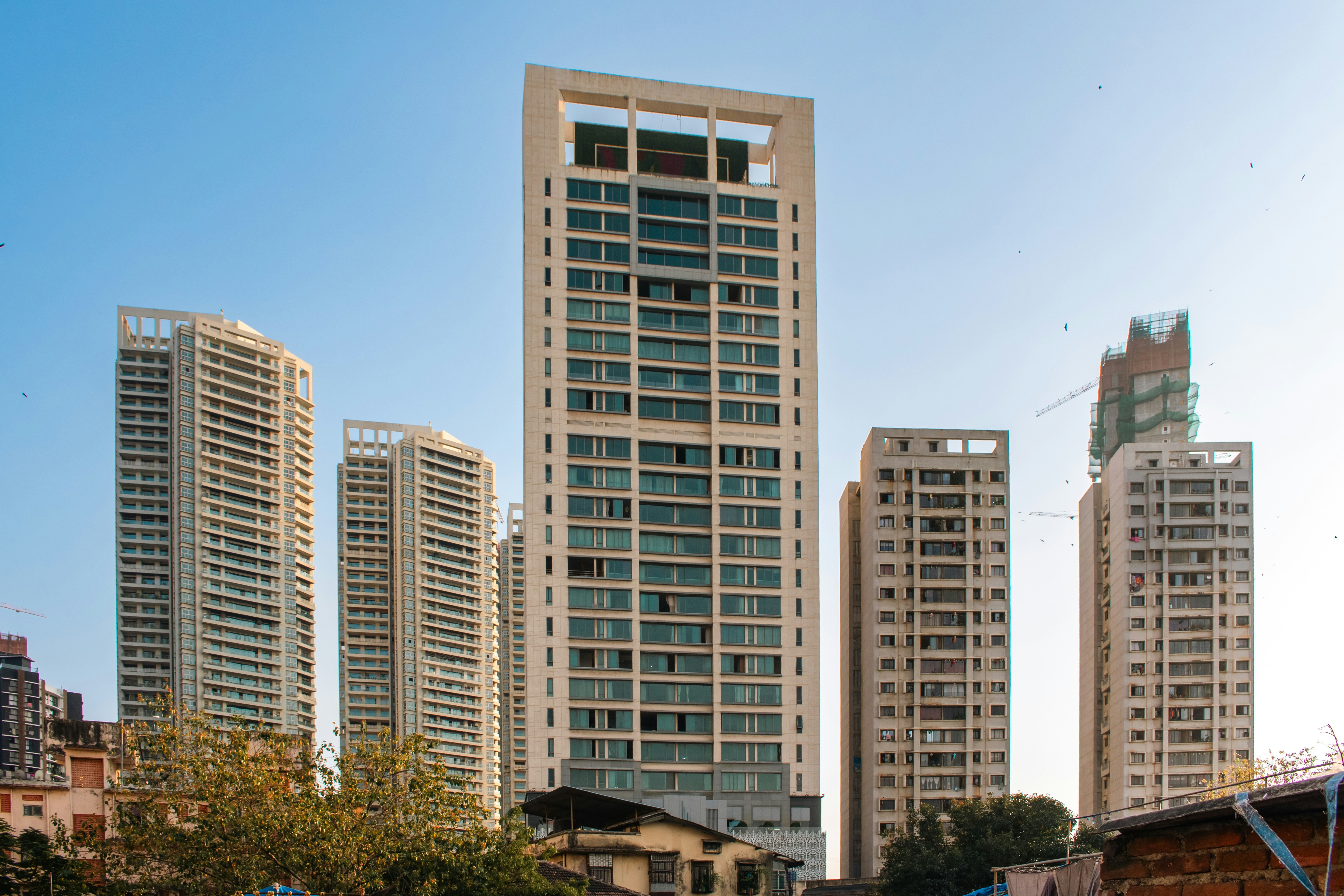 Several modern, towering high-rise residential buildings in Mumbai, dominating the skyline under a bright, clear sky. In sharp contrast, the immediate foreground shows the makeshift, low-rise structures of the Dhobi Ghat settlement, including a small section of roof and trees. The disparity in scale and architecture between the sprawling towers and the humble shanties below powerfully illustrates the extreme urban contrast and socio-economic stratification characteristic of the city. This is highly suitable for content on Indian urbanization, social inequality, and cityscape photography.