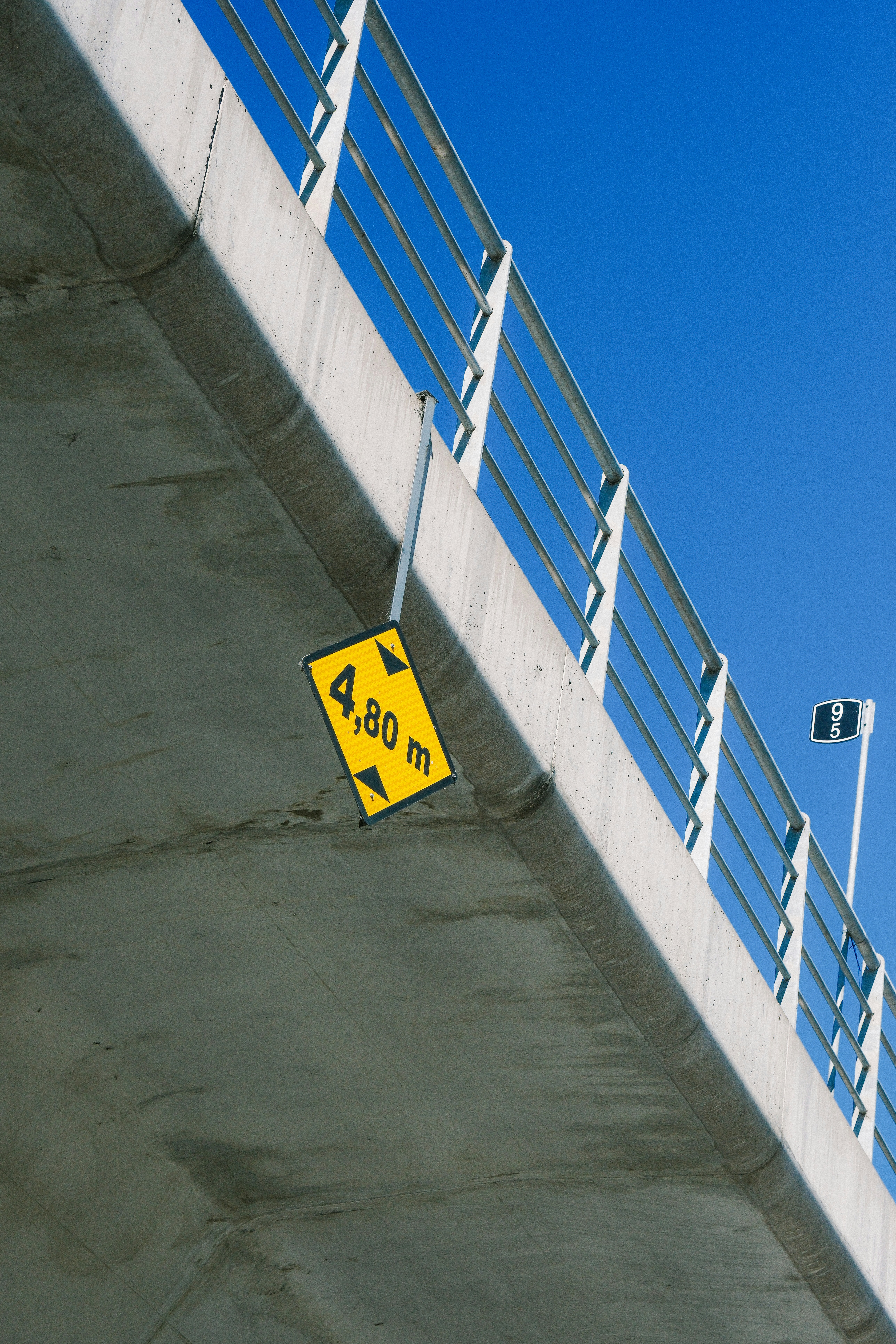 Tilted Yellow Clearance Sign Under a Sunlit Concrete Overpass