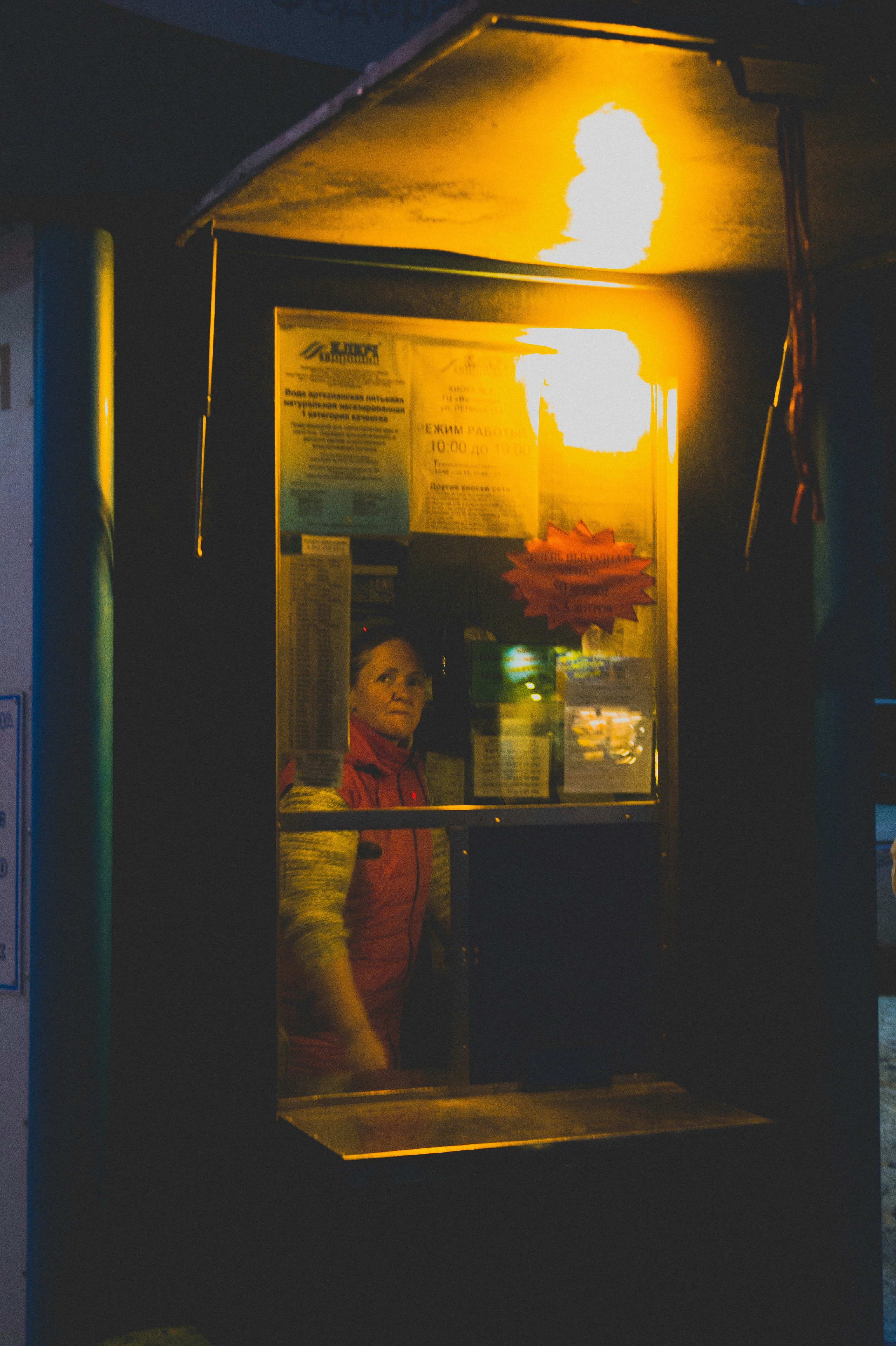 A woman looks out from a small, brightly lit booth at night, her face illuminated by the warm overhead light in a dark urban setting.