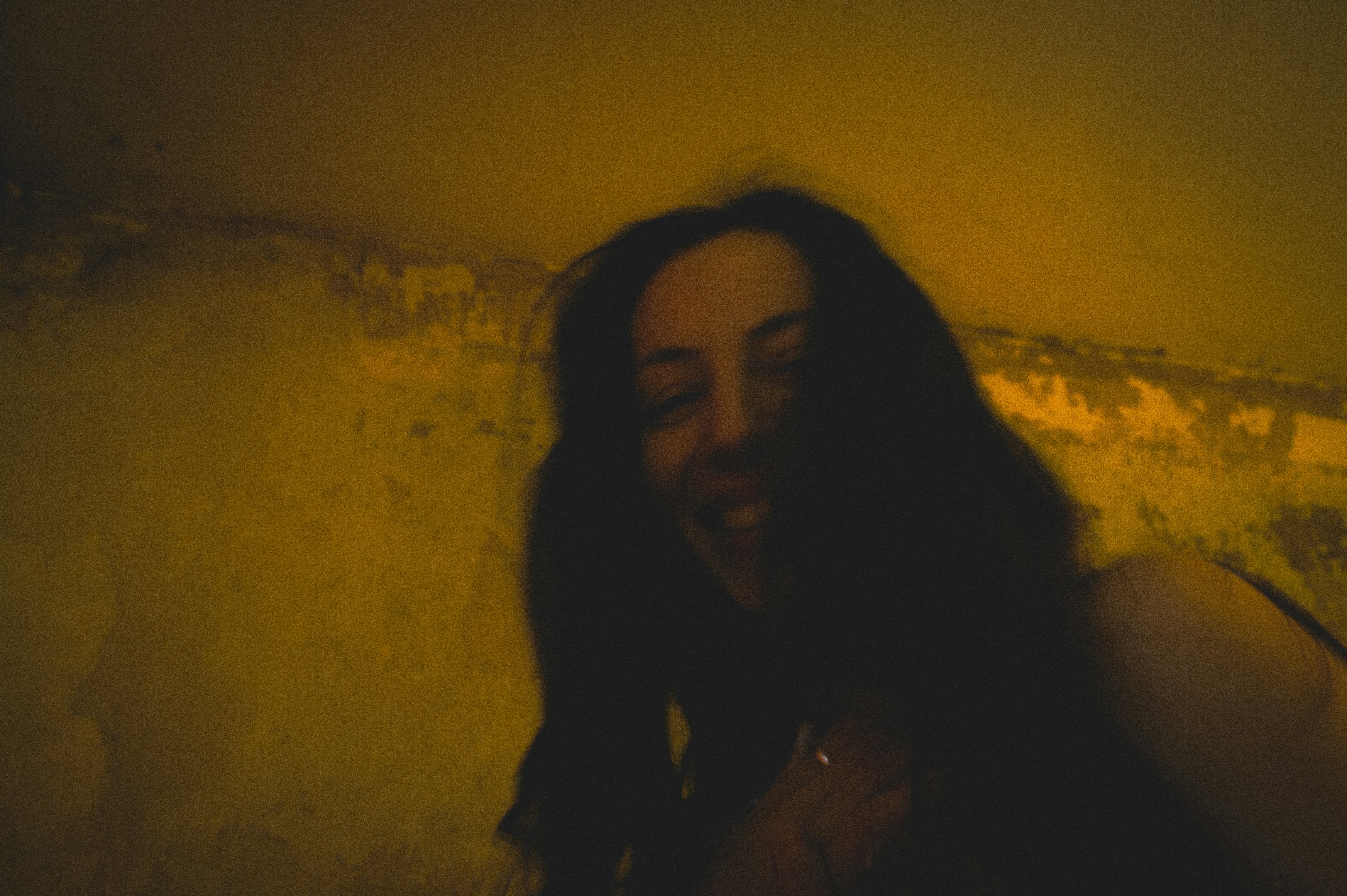 A blurred, low-light portrait captures the spontaneous smile of a woman with long dark hair, bathed in a warm, yellowish glow.