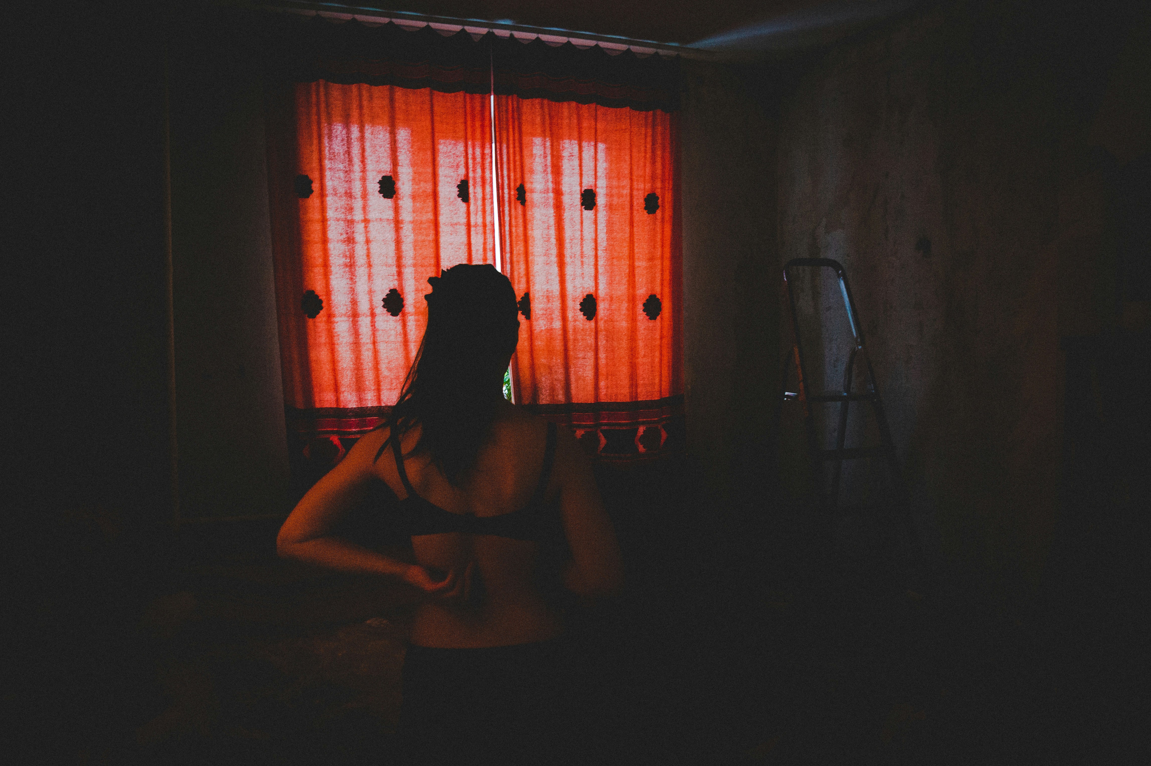 A woman seen from behind, adjusting her bra in a dimly lit room, with an orange curtain-covered window as a backdrop.