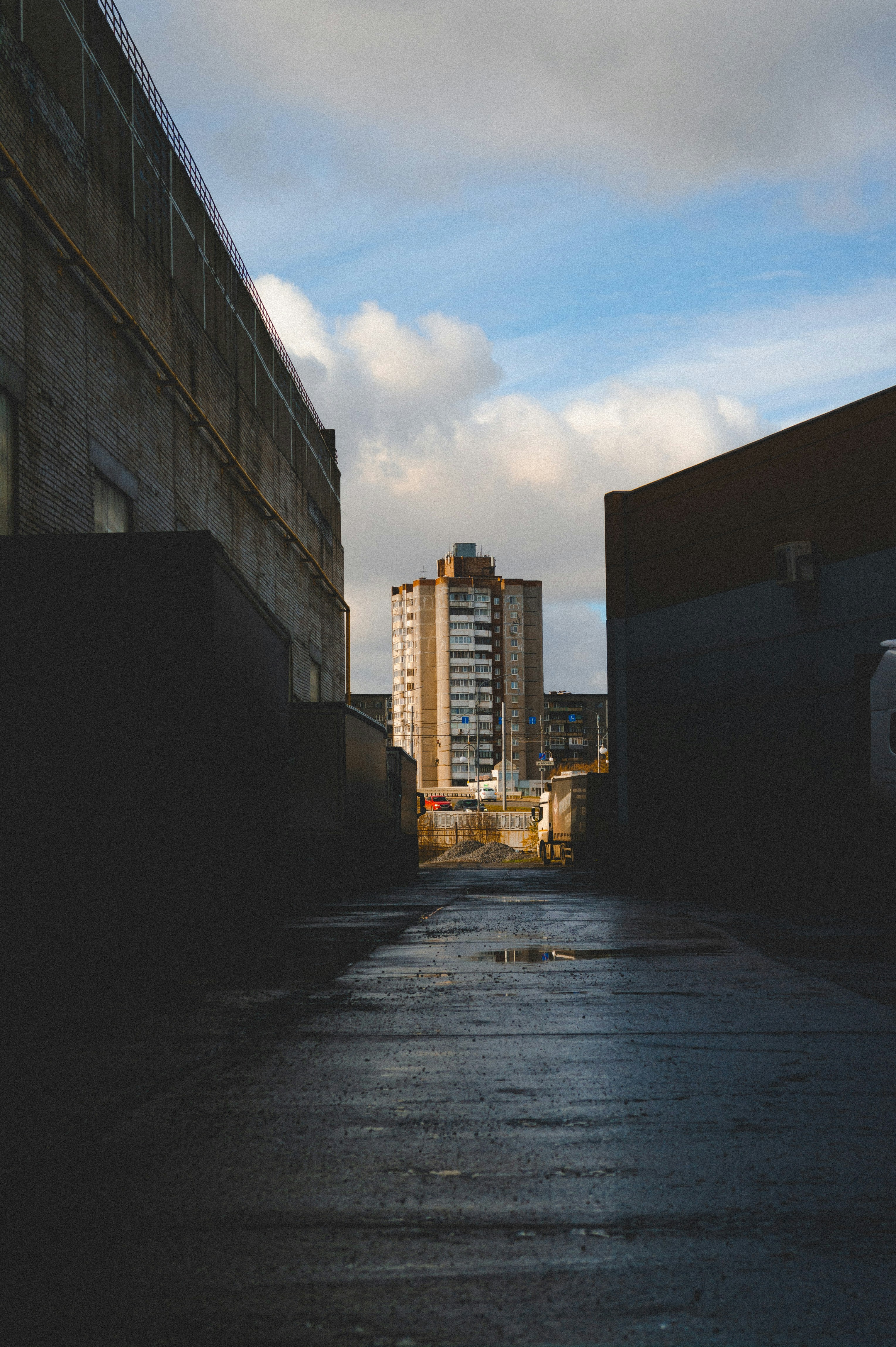 A lone high-rise building stands at the end of a dark, wet urban alleyway, catching the light under a cloudy sky.