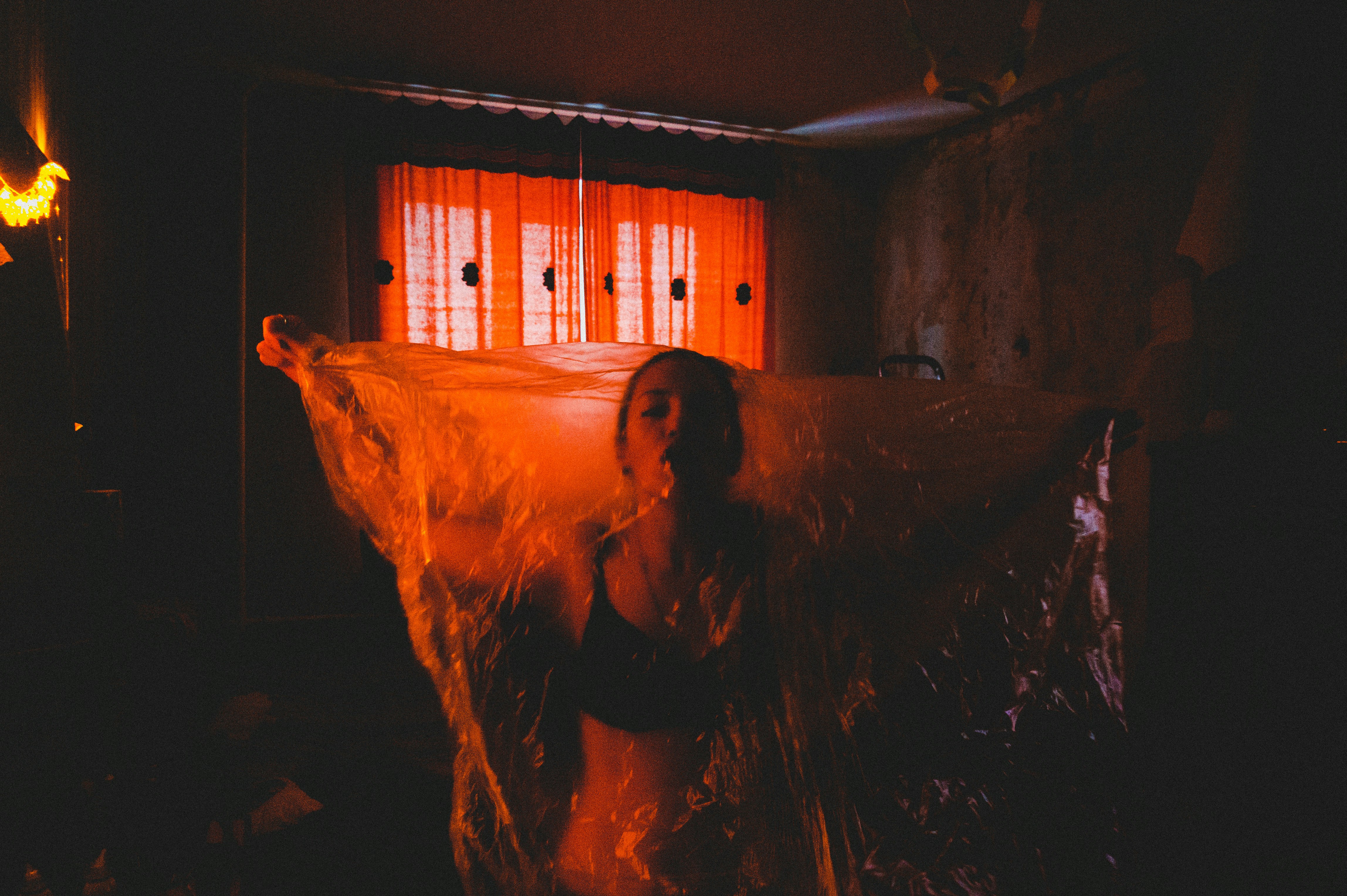 A mysterious portrait of a woman enveloped in plastic, illuminated by a warm orange glow from a window in a dimly lit room.