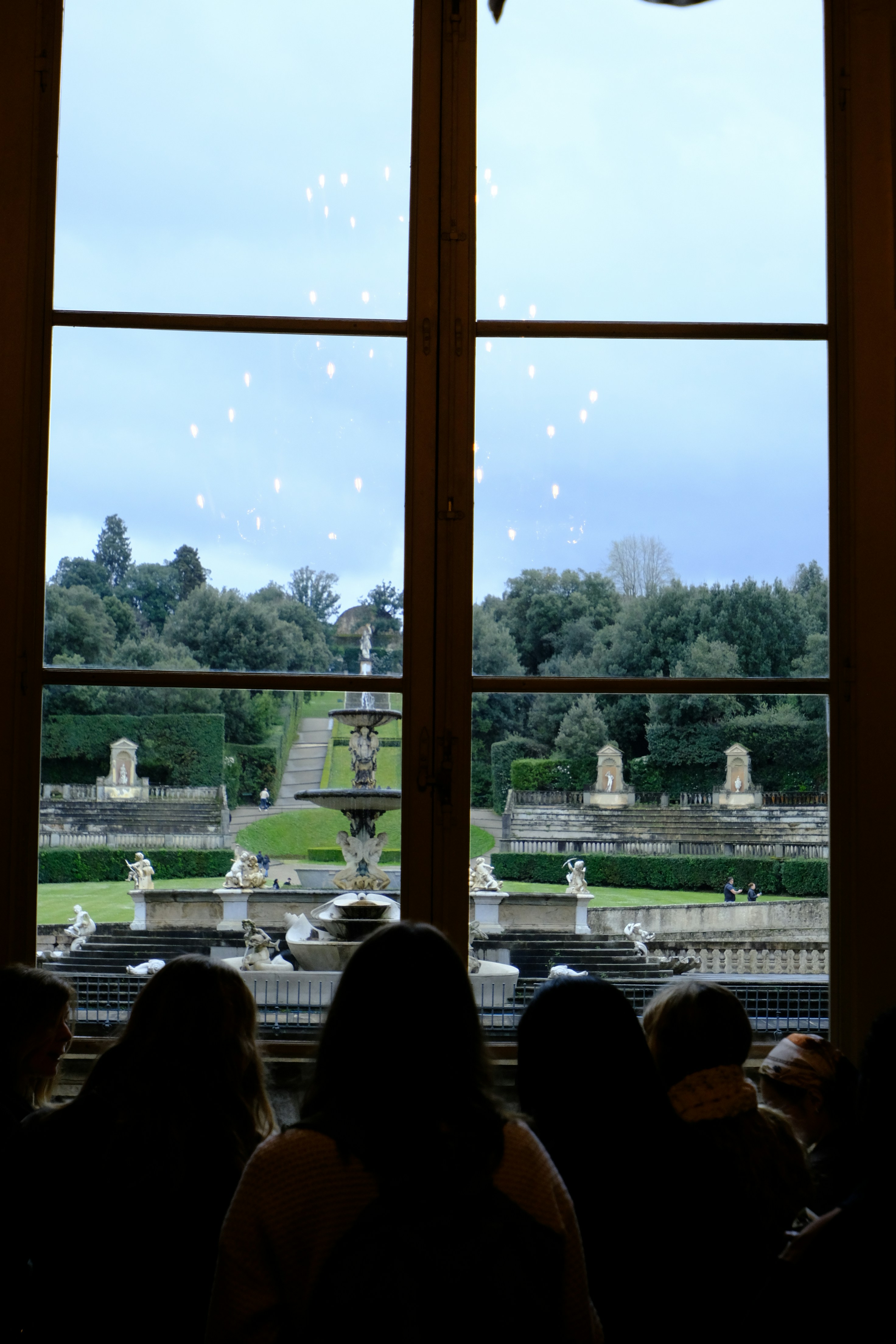 Silhouetted Visitors Gaze Through a Grand Window at a Formal Garden Fountain