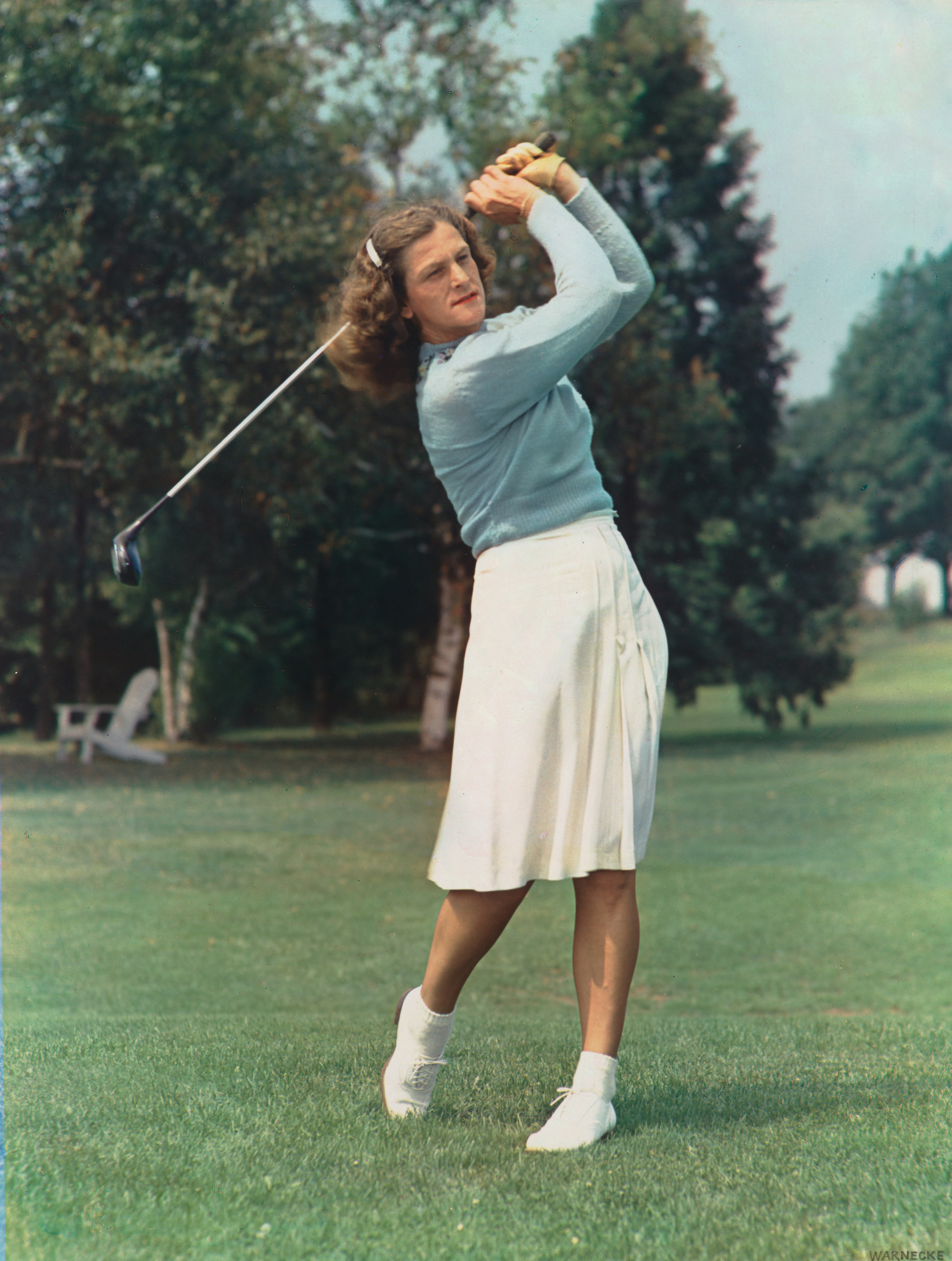 Woman mid-swing on a sunny golf course, attire suggests vintage 1970s athletic wear; outdoor green backdrop and a club in motion.
