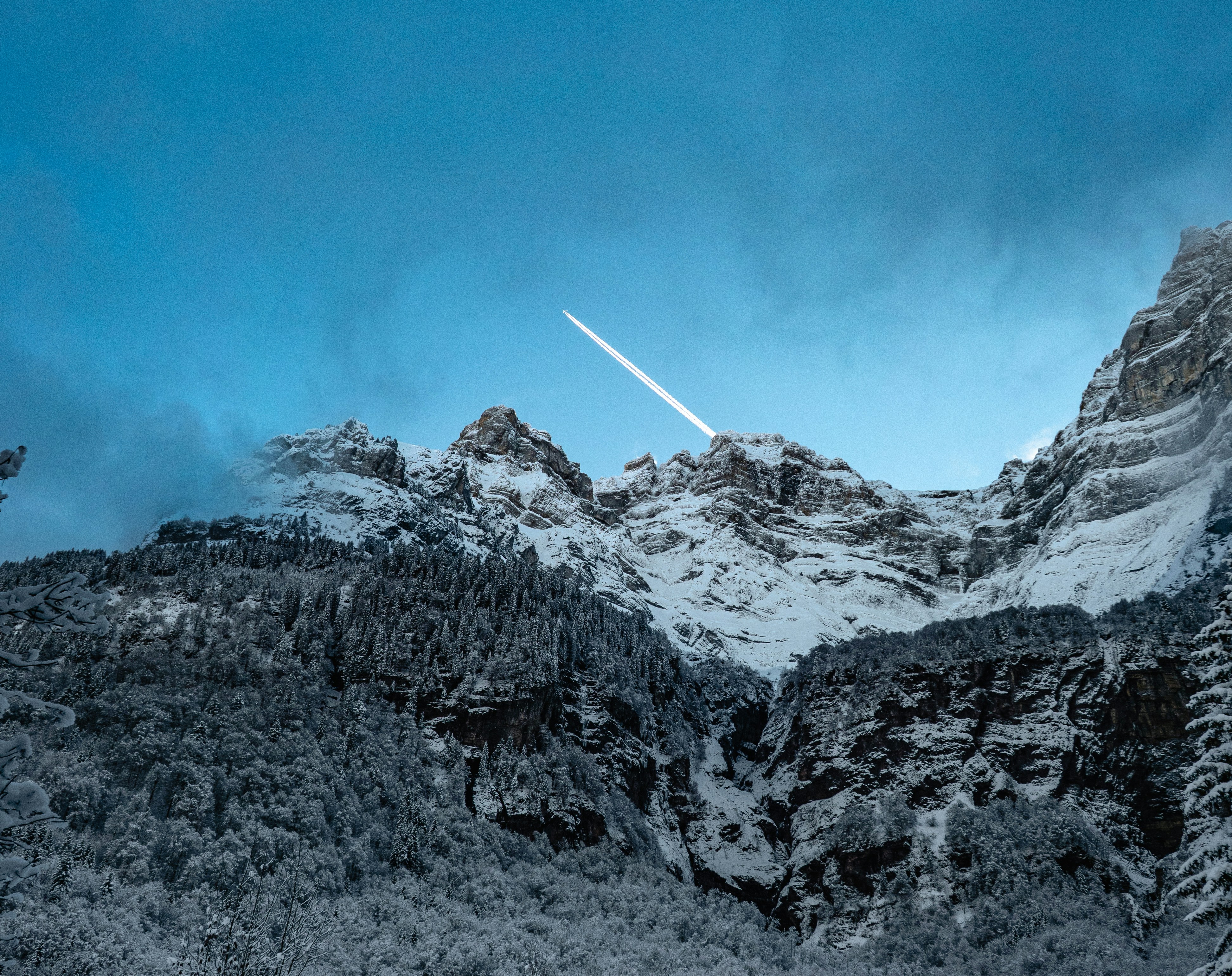 White contrail cutting through alpine dusk above snow-kissed peaks