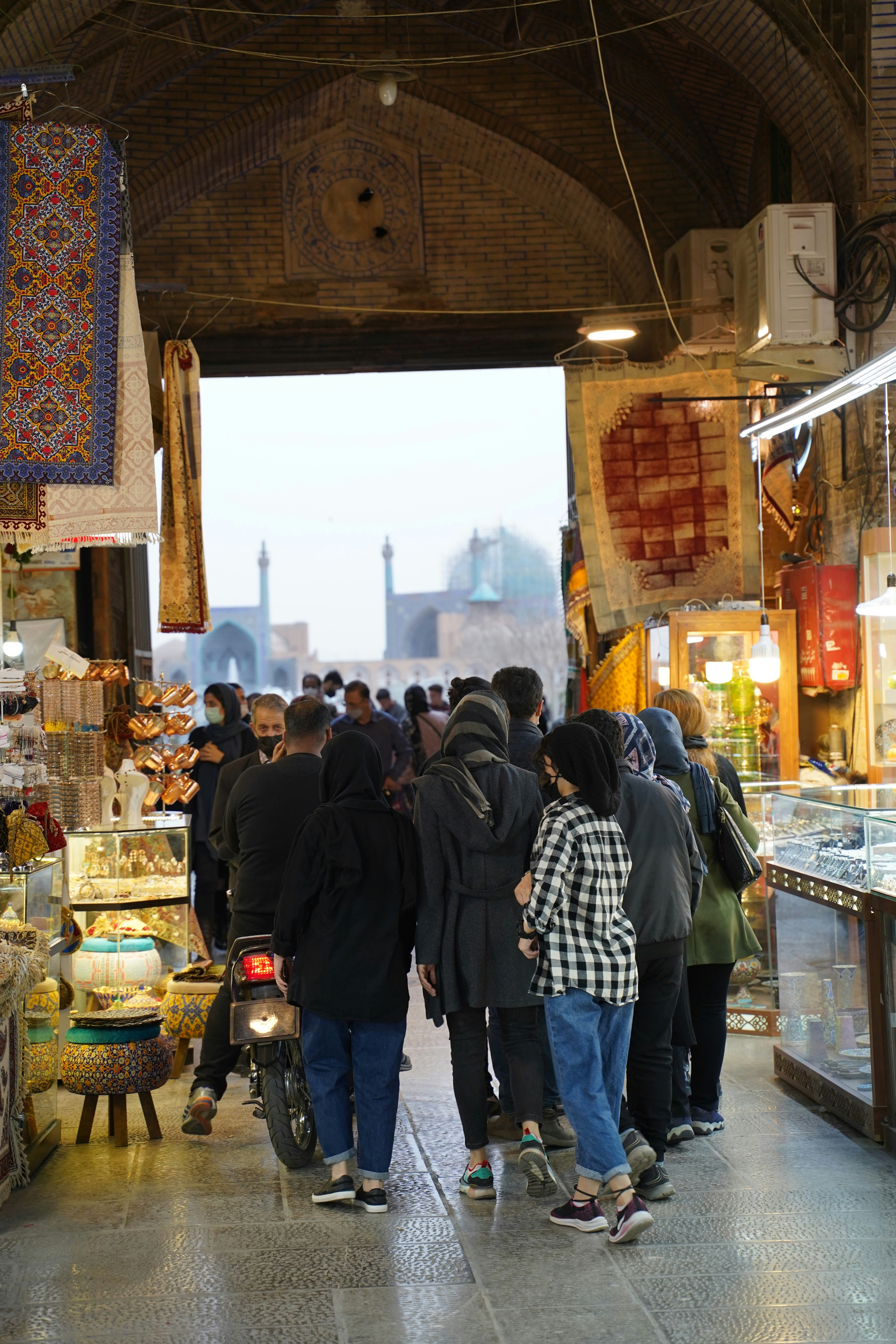 A lively scene inside the historic Qeysarieh Bazaar of Naqsh-e Jahan Square, where people walk past colorful handicrafts, hanging carpets, and glowing shop displays under the old brick archways.