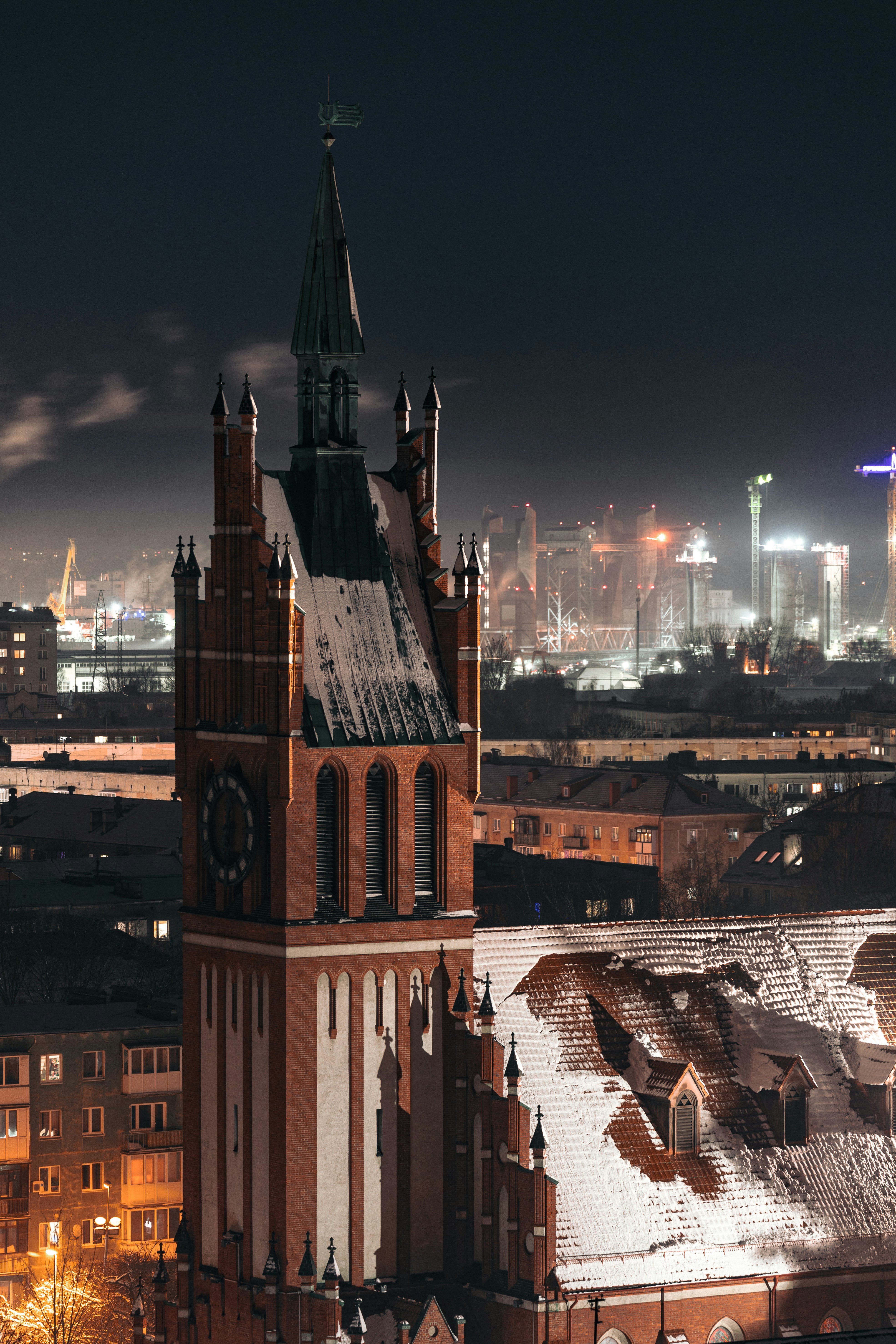 Kaliningrad Regional Philharmonic Hall on a snowy winter night, captured from above with the neo‑Gothic brick tower in the foreground and illuminated industrial cranes and factories fading into the misty background. Soft warm light from the city windows contrasts with the cold blue night sky and snow-covered roof tiles, creating a cinematic urban landscape in Russia’s westernmost city.
