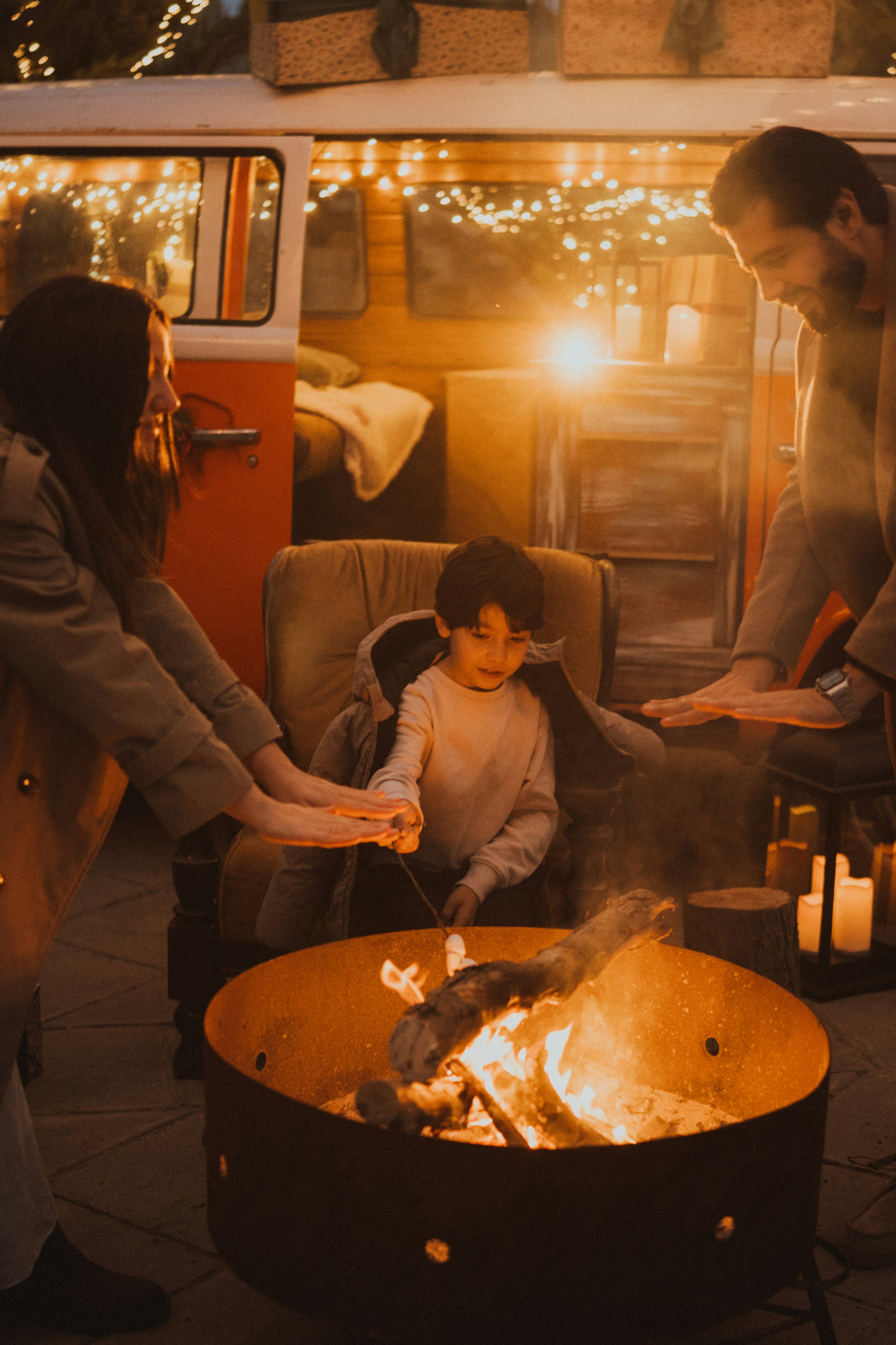 Warm campfire scene inside a vintage van with a family gathered around the flames; a child sits in a chair reaching toward the fire while adults nearby help and smile. This is a candid lifestyle photo, not a logo or illustration.