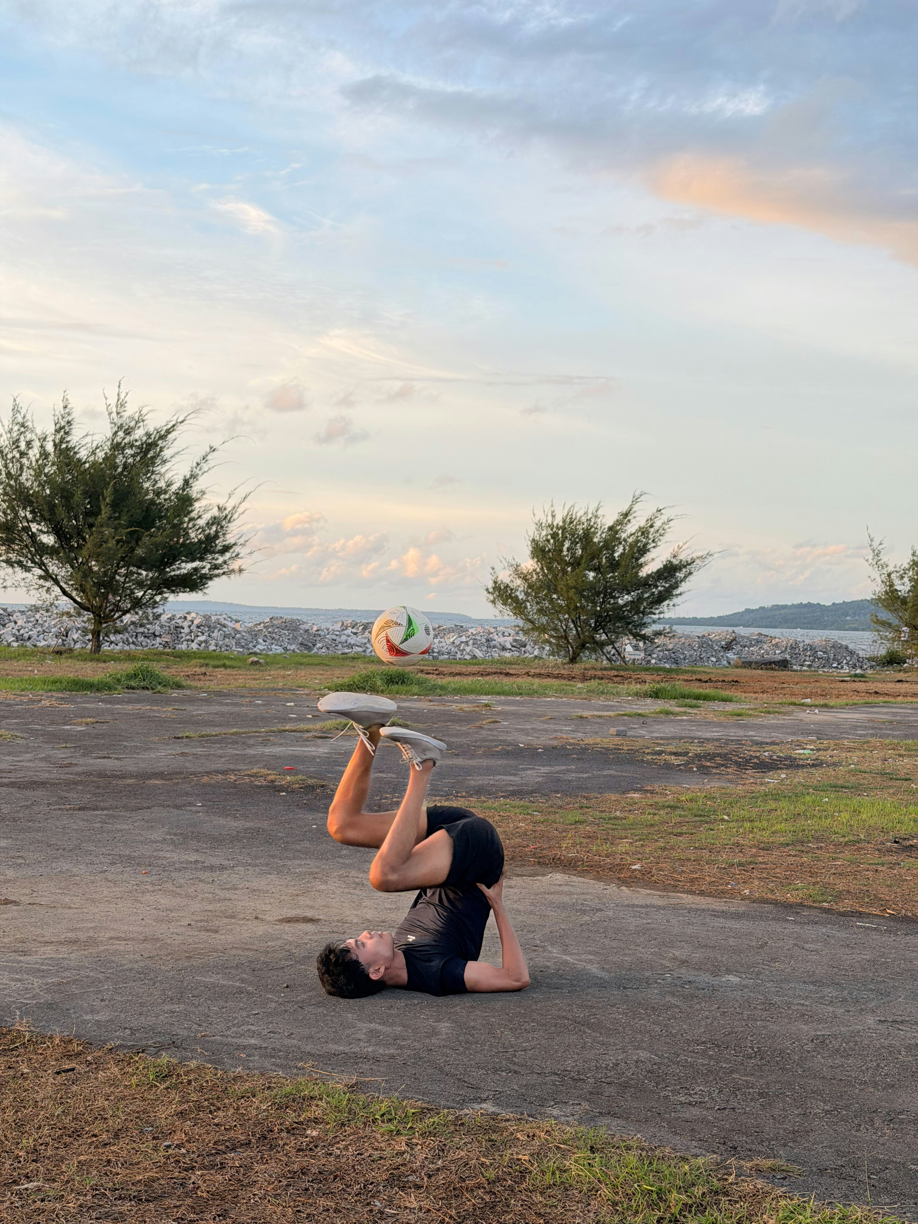 Young athlete balances feet high in the air over a flat court with a volleyball, relaxed background trees and water beyond.