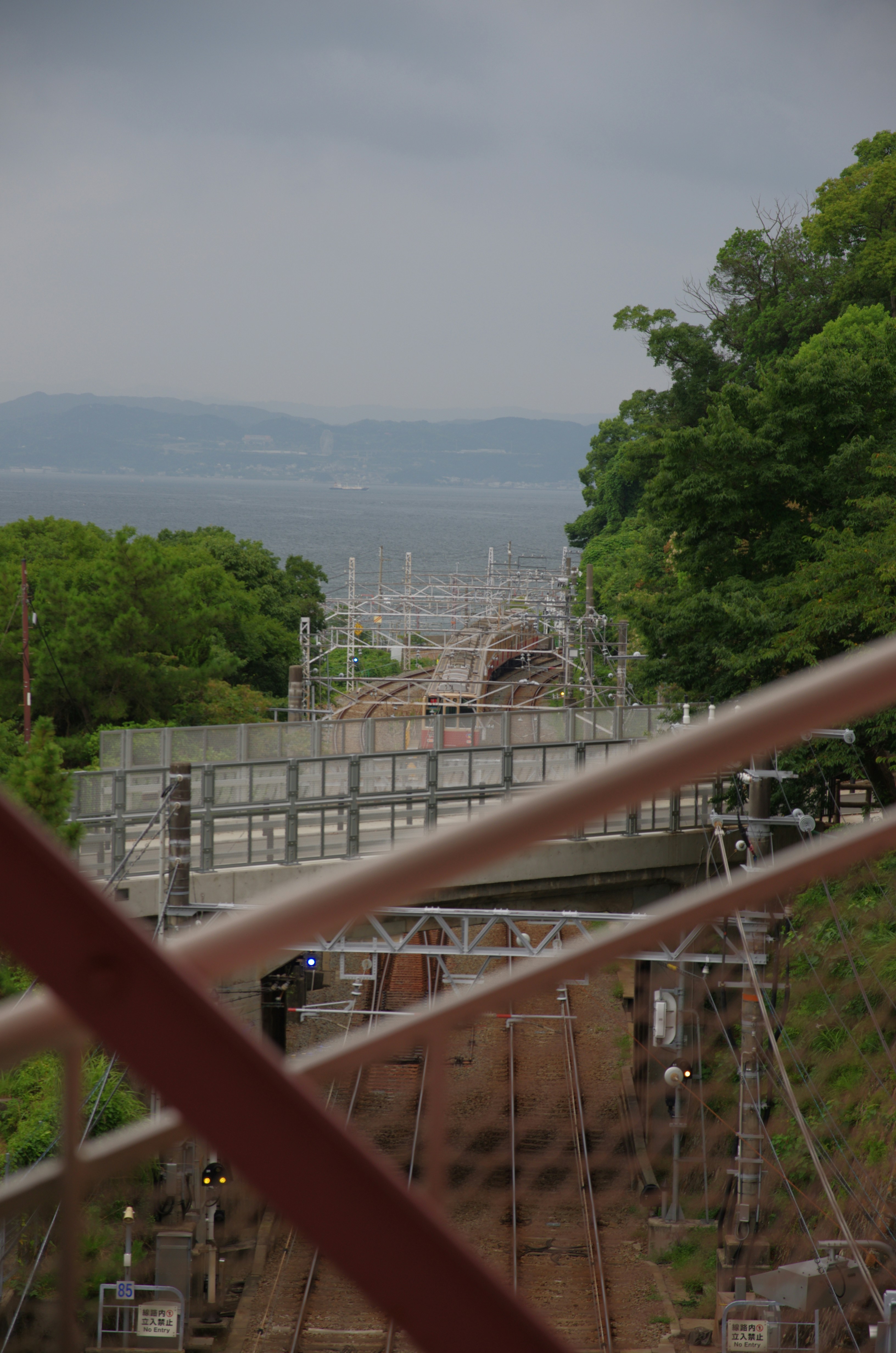 Railway tracks and a tunnel under construction framed by green trees with a view toward the sea in the distance. A diagonal railing in the foreground suggests a viewpoint from a bridge or platform.