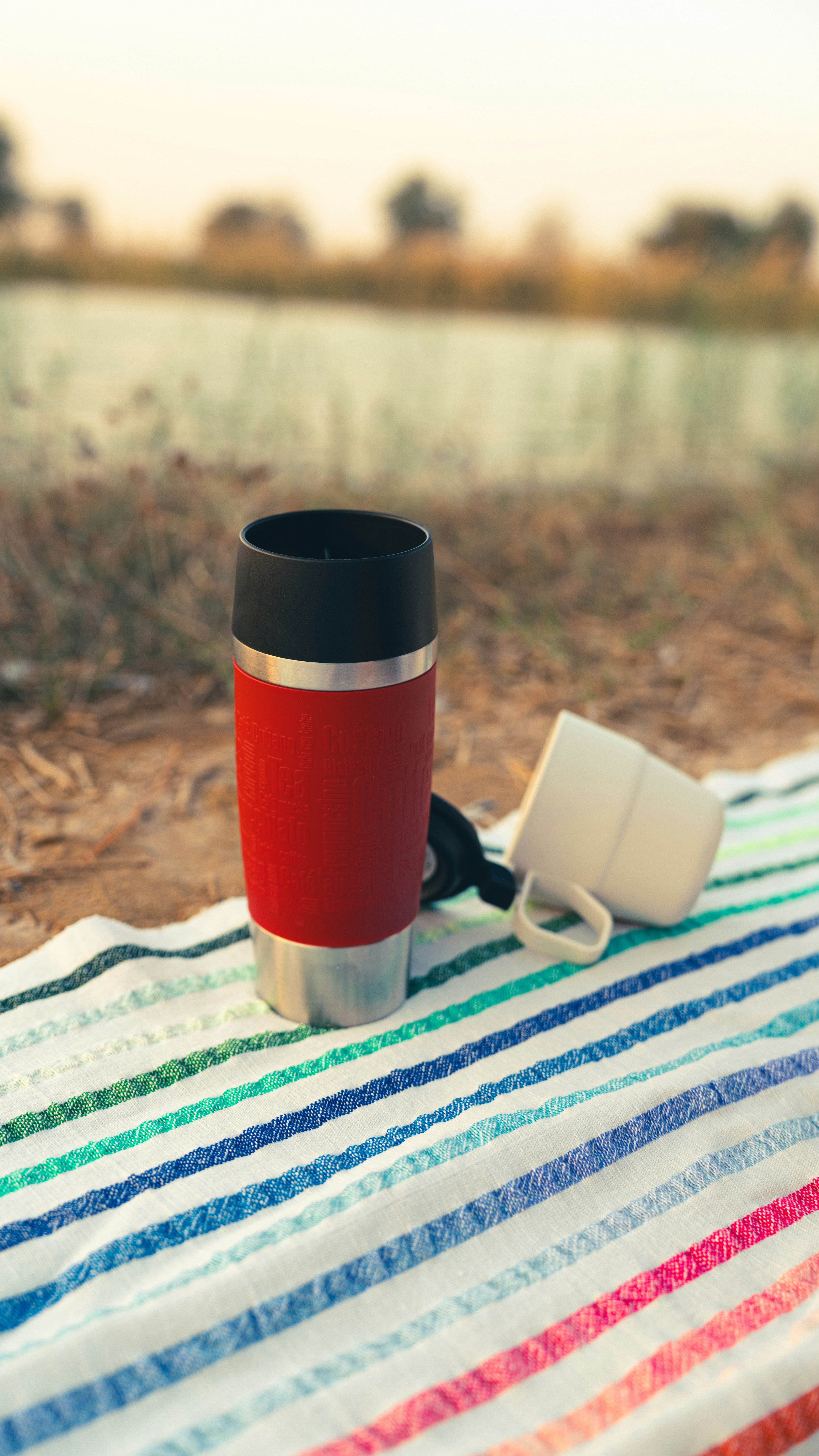 Hydrating Moment: A Stainless Steel Thermos rests on a striped towel near a calm lakeshore at golden hour
