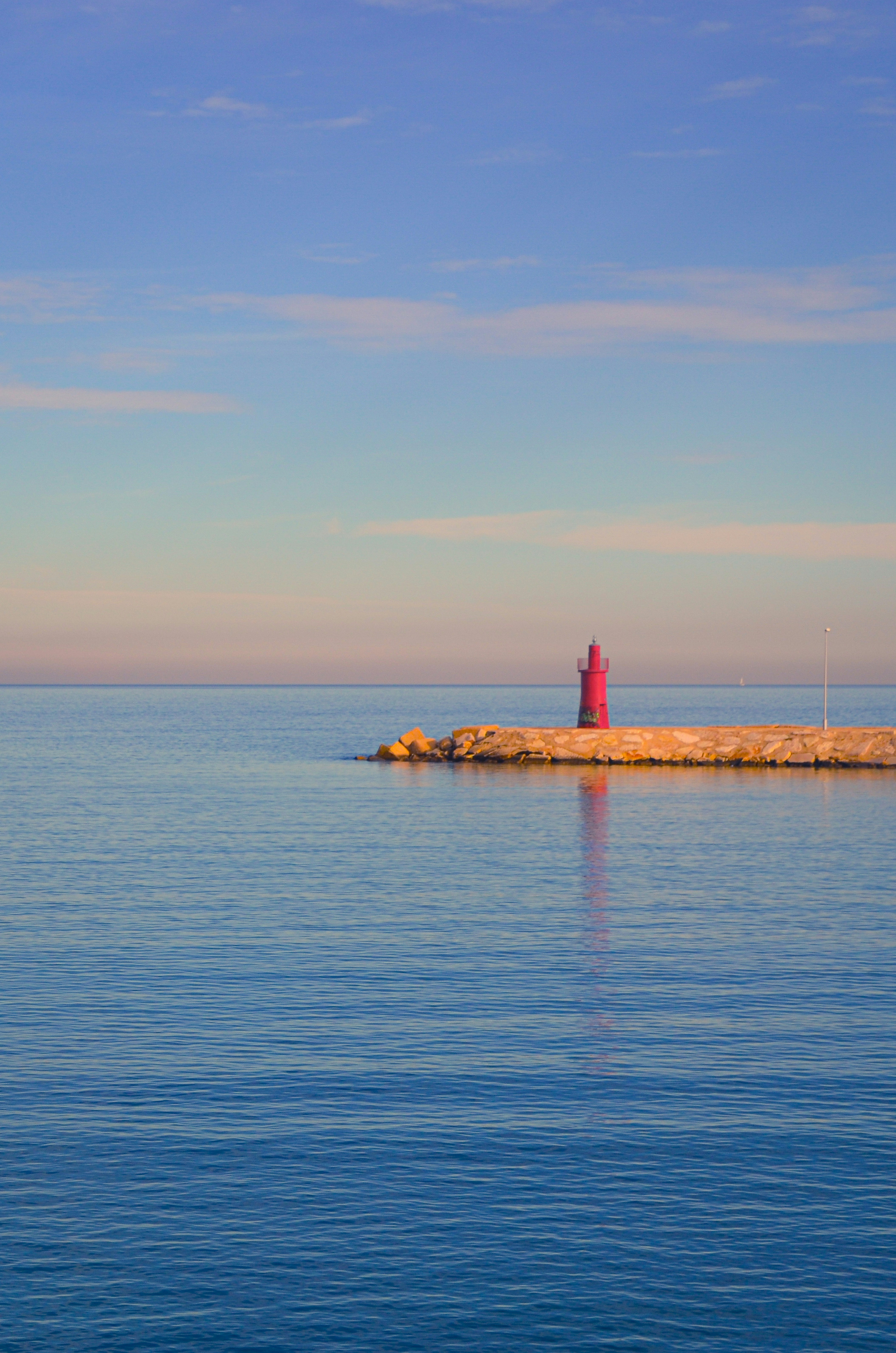 A red lighthouse stands on a rocky pier, reflecting in calm waters under a pastel sunset sky. The maritime scene blends functionality with beauty, offering a tranquil moment by the sea.