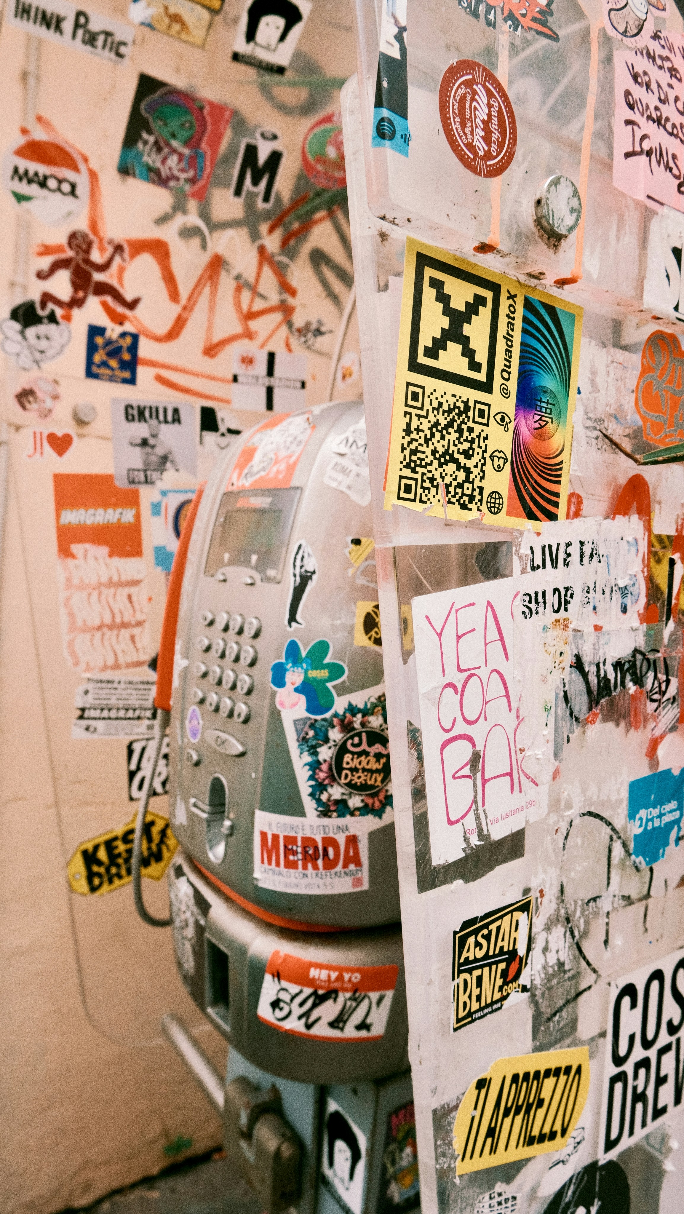 A public payphone covered in colorful street art and stickers, photographed in an urban alley with a gritty, creative atmosphere