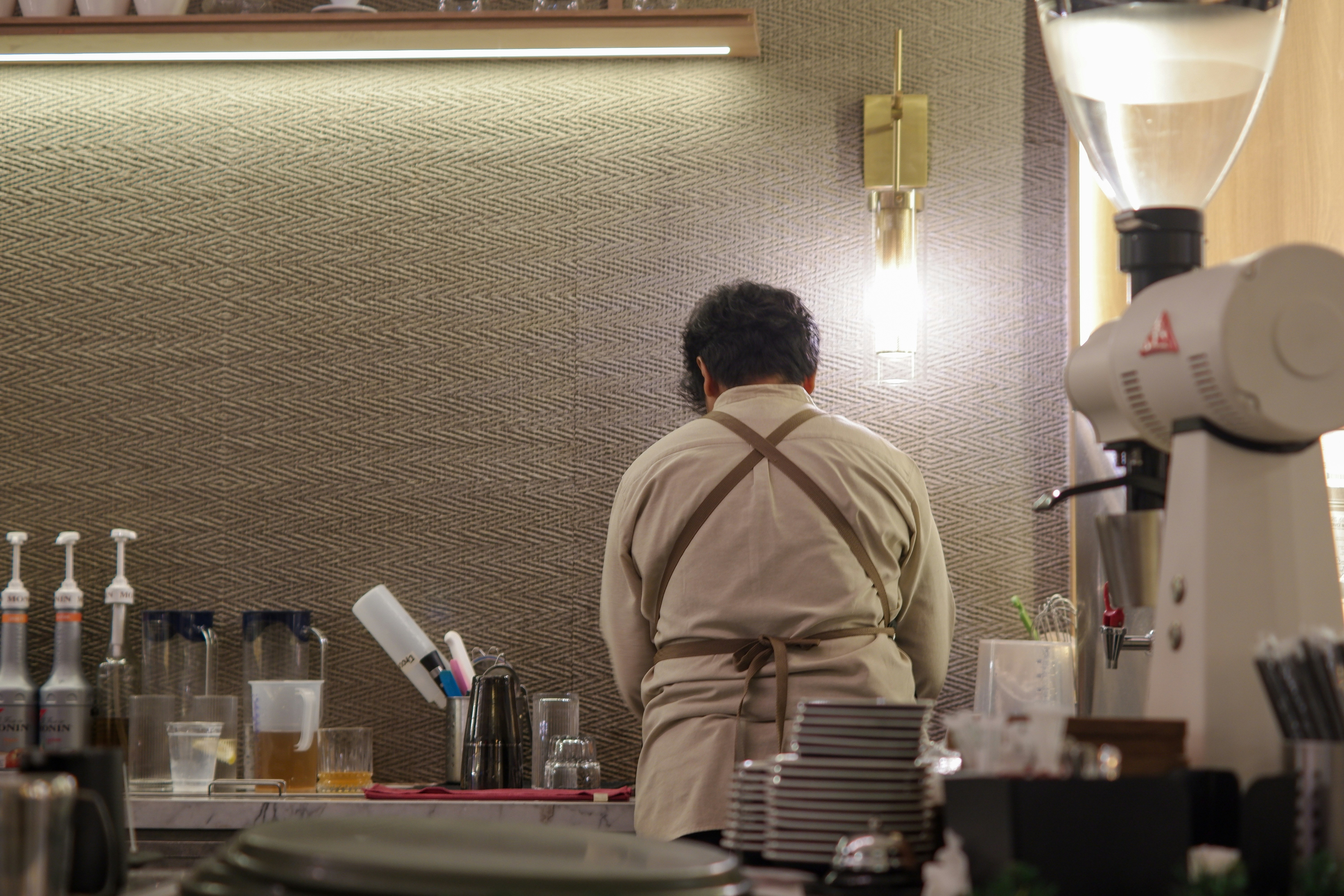 A quiet moment behind the counter as the barista prepares drinks, framed by warm café lighting and soft textures.