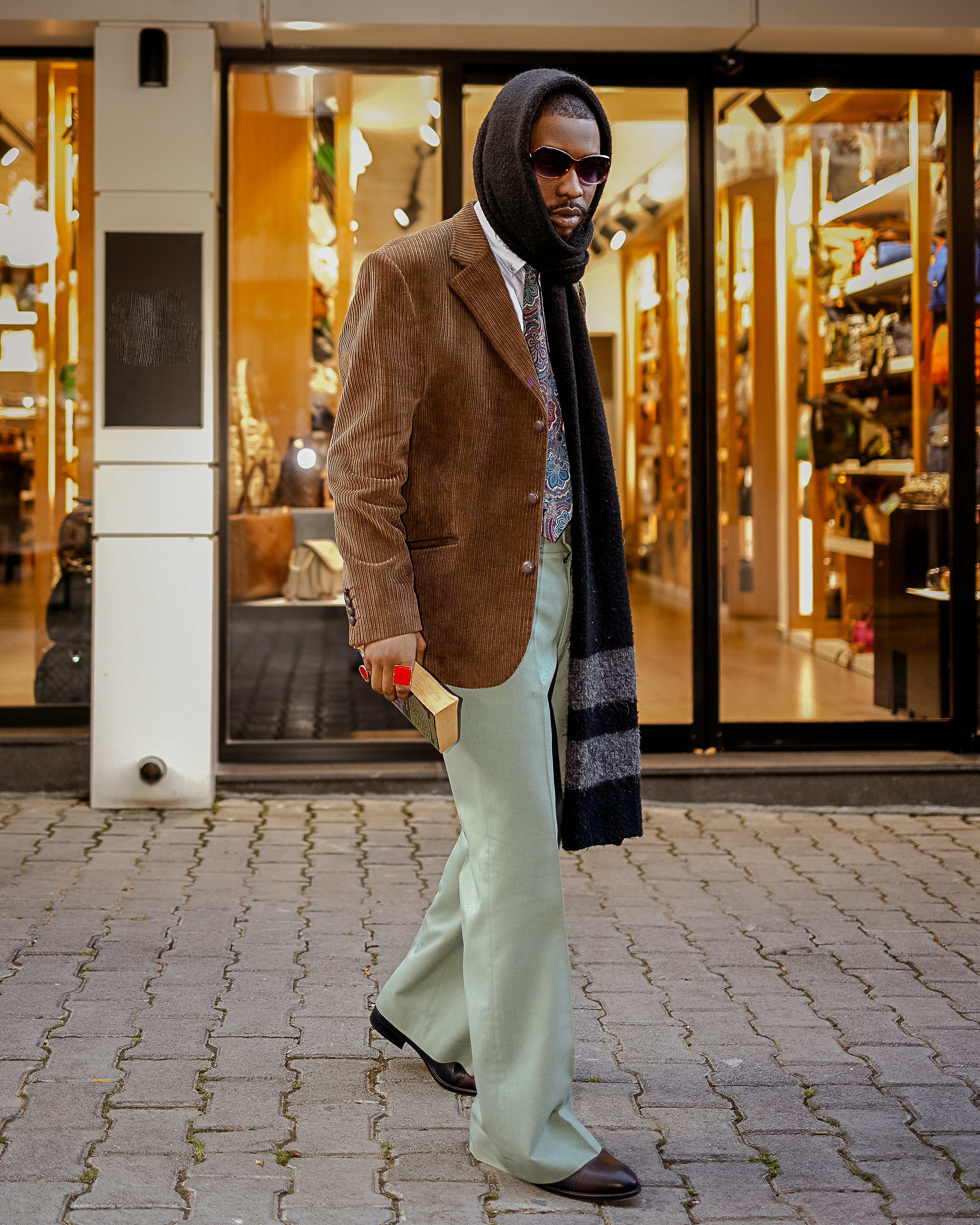 A portrait of a confident, stylish man posing in an urban setting. He is wearing a brown corduroy jacket, a colorful paisley tie, and dark sunglasses, accessorized with statement rings and a scarf.