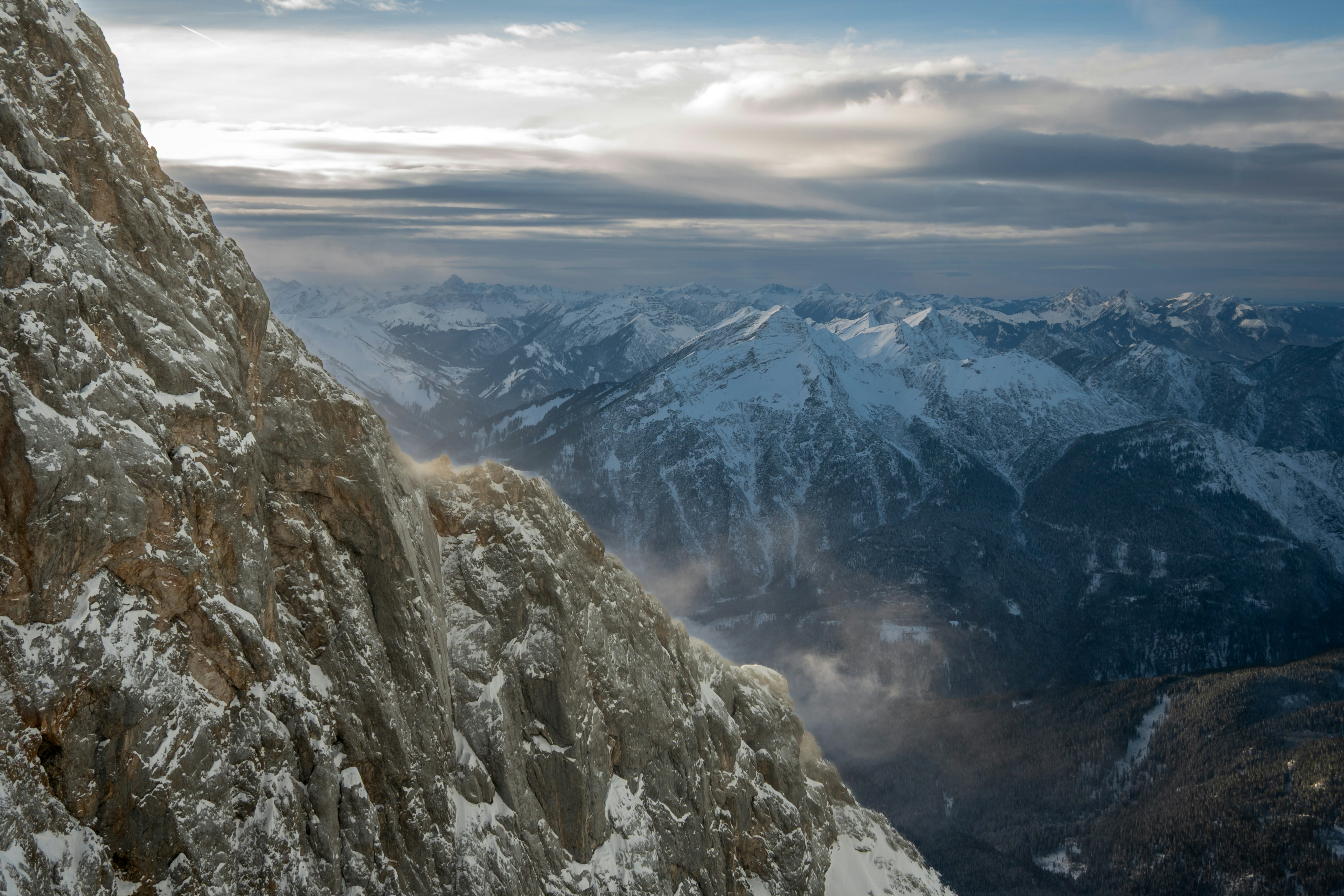 Snow-dusted cliffside with distant Alpine ridges under a dramatic cloudscape