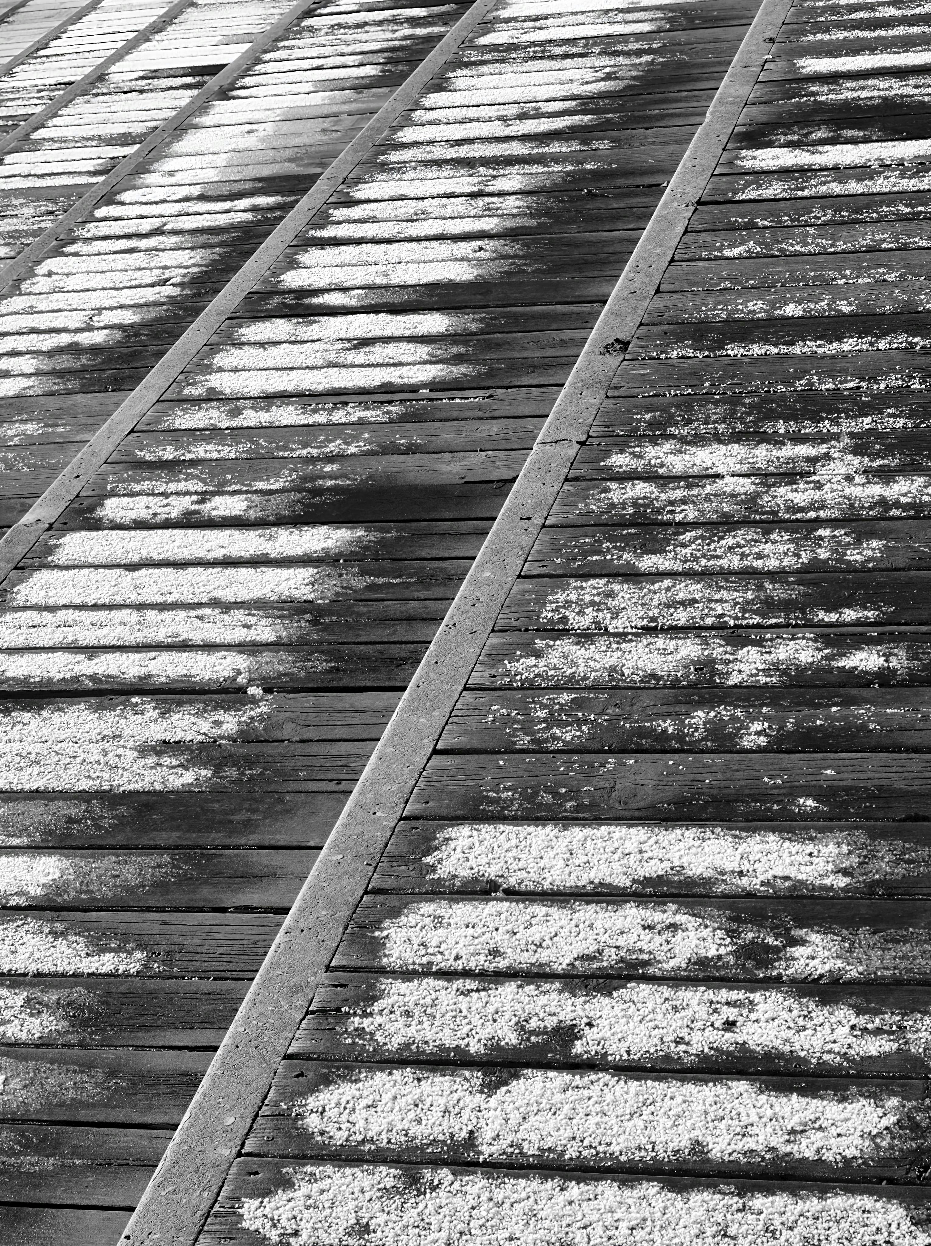 A high-contrast black and white abstract photograph focusing intensely on texture and linear perspective. The image captures a section of a wooden deck or walkway—possibly a pier or set of stairs—where snow lies in uneven, horizontal stripes. The dark wooden planks provide a deep, textured background, while the white snow bands rhythmically divide the space. A strong diagonal line, likely a metal or concrete rail, cuts across the frame, enhancing the geometric complexity and dynamic movement of the composition. This piece functions as a study in pattern, rhythm, and the elegant contrast betwee