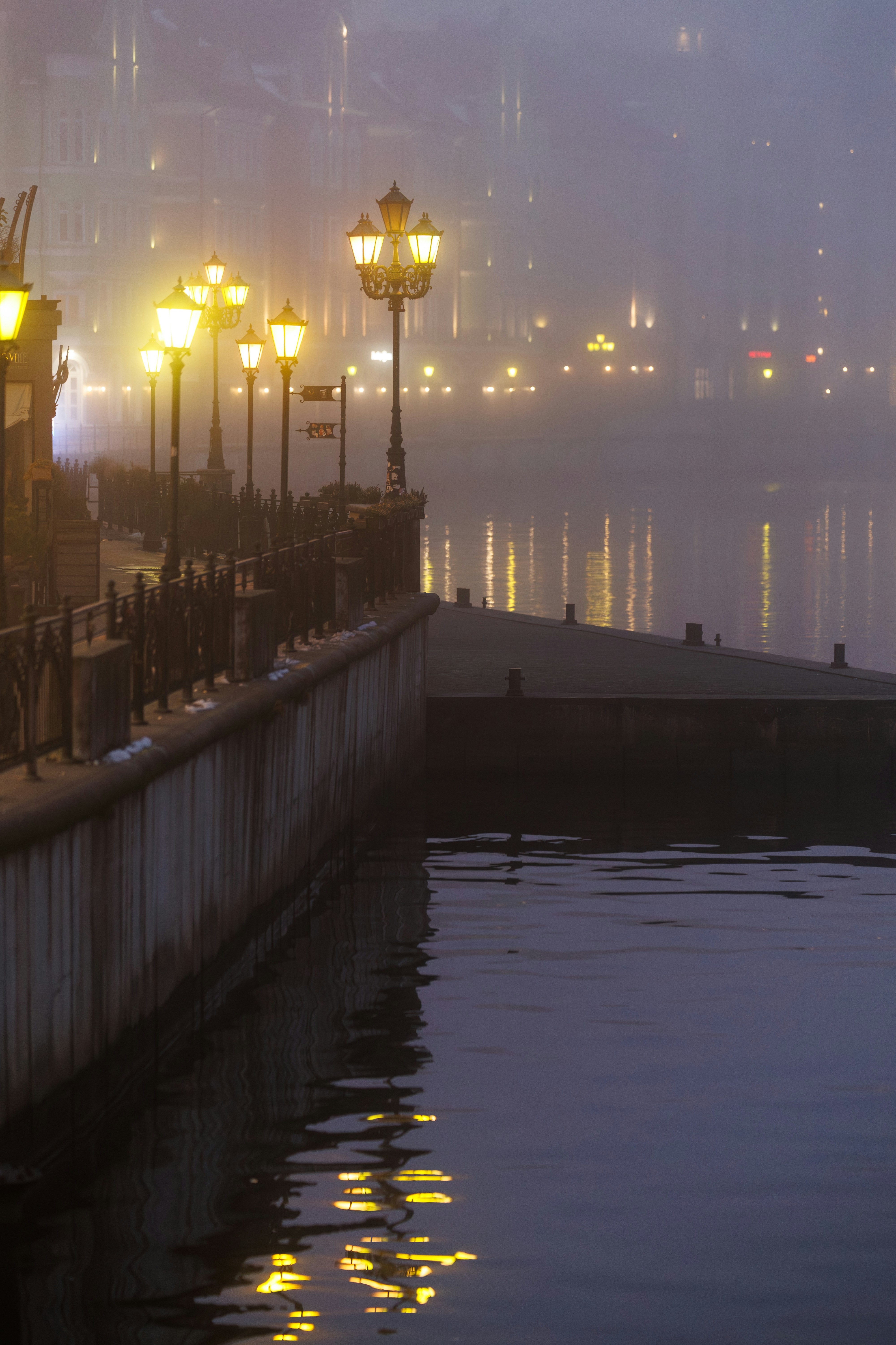 A row of glowing street lamps dissolves into thick fog along the waterfront, their reflections shimmering softly on the river.