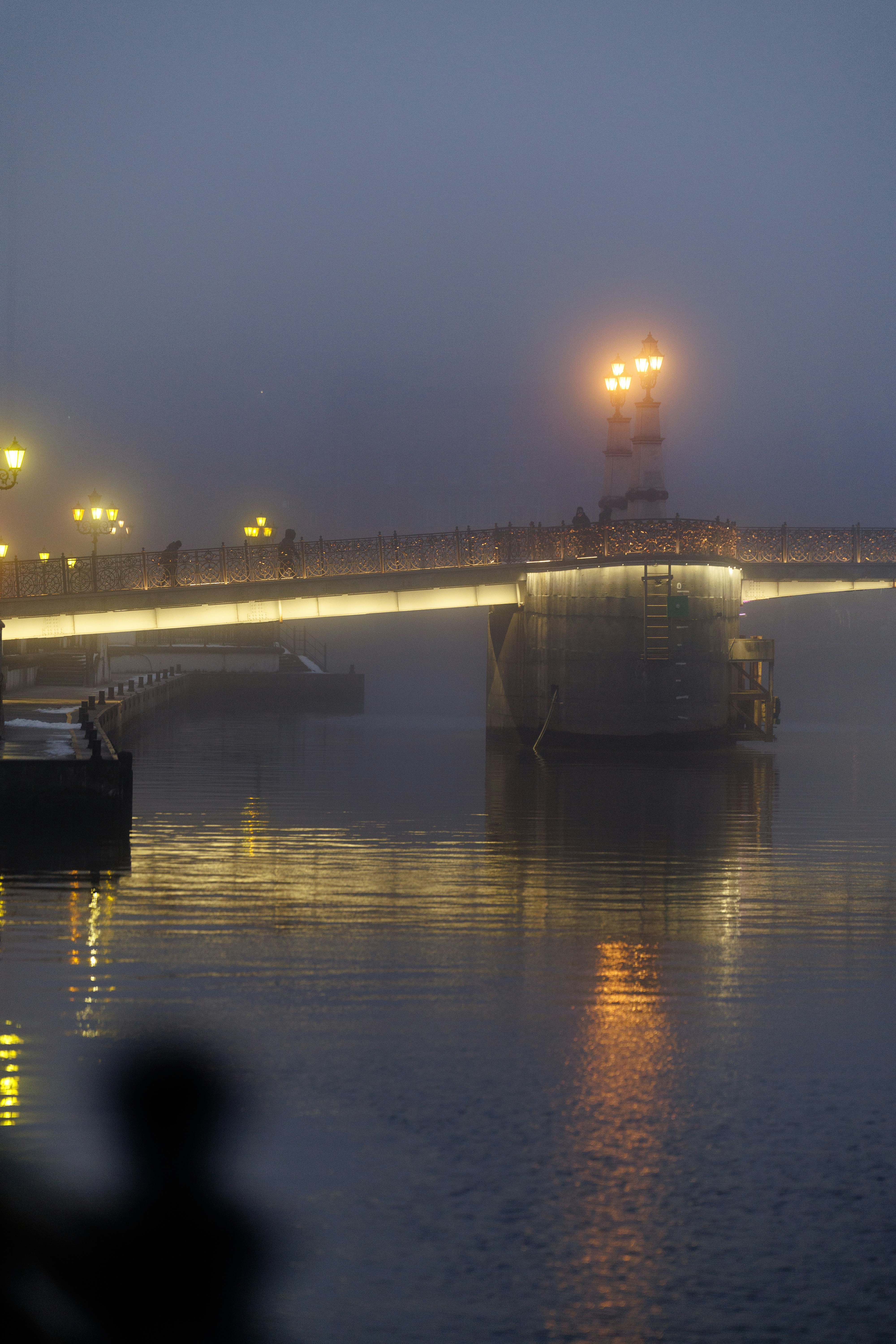 A long, softly lit bridge fades into a heavy fog, its warm lights reflecting gently across the quiet river.