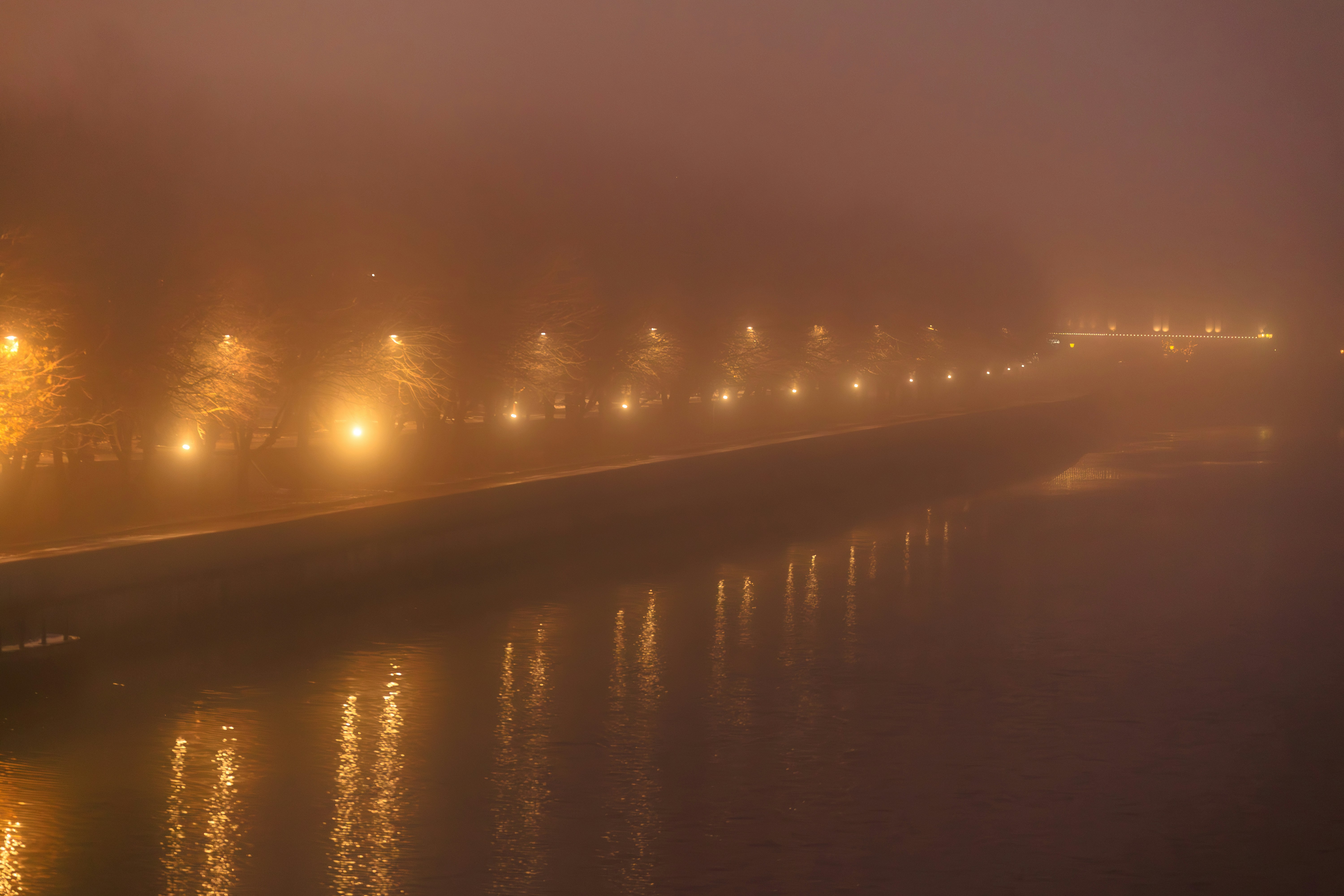A glowing line of street lamps stretches along a fog-filled riverside path, their warm reflections fading gently into the misty distance.