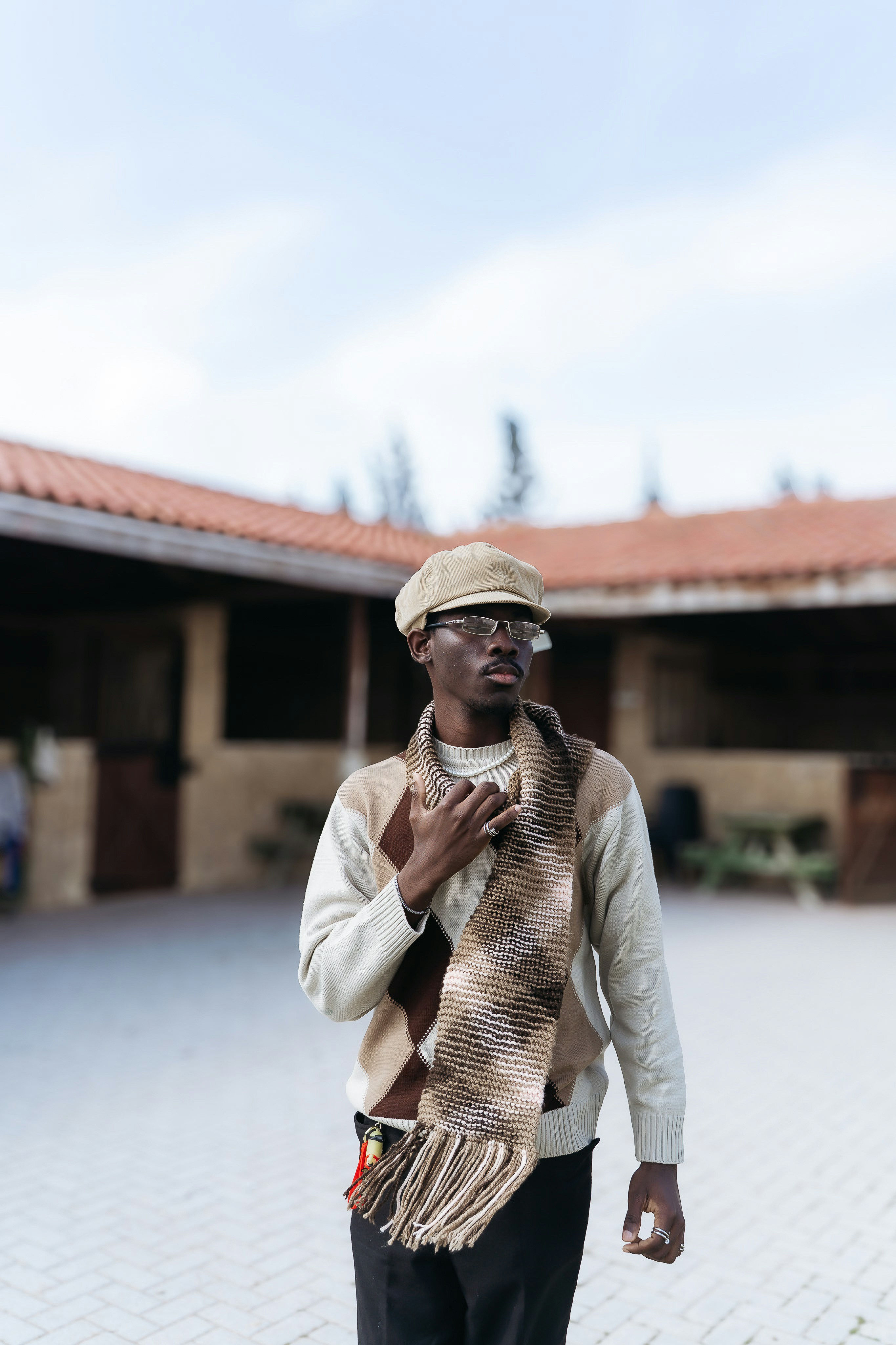 A close-up outdoor portrait featuring a person in vintage-inspired fashion, including a neutral-toned sweater, textured scarf, and cap. Captured with natural light and a soft background, the image highlights modern style mixed with classic, earthy colors.